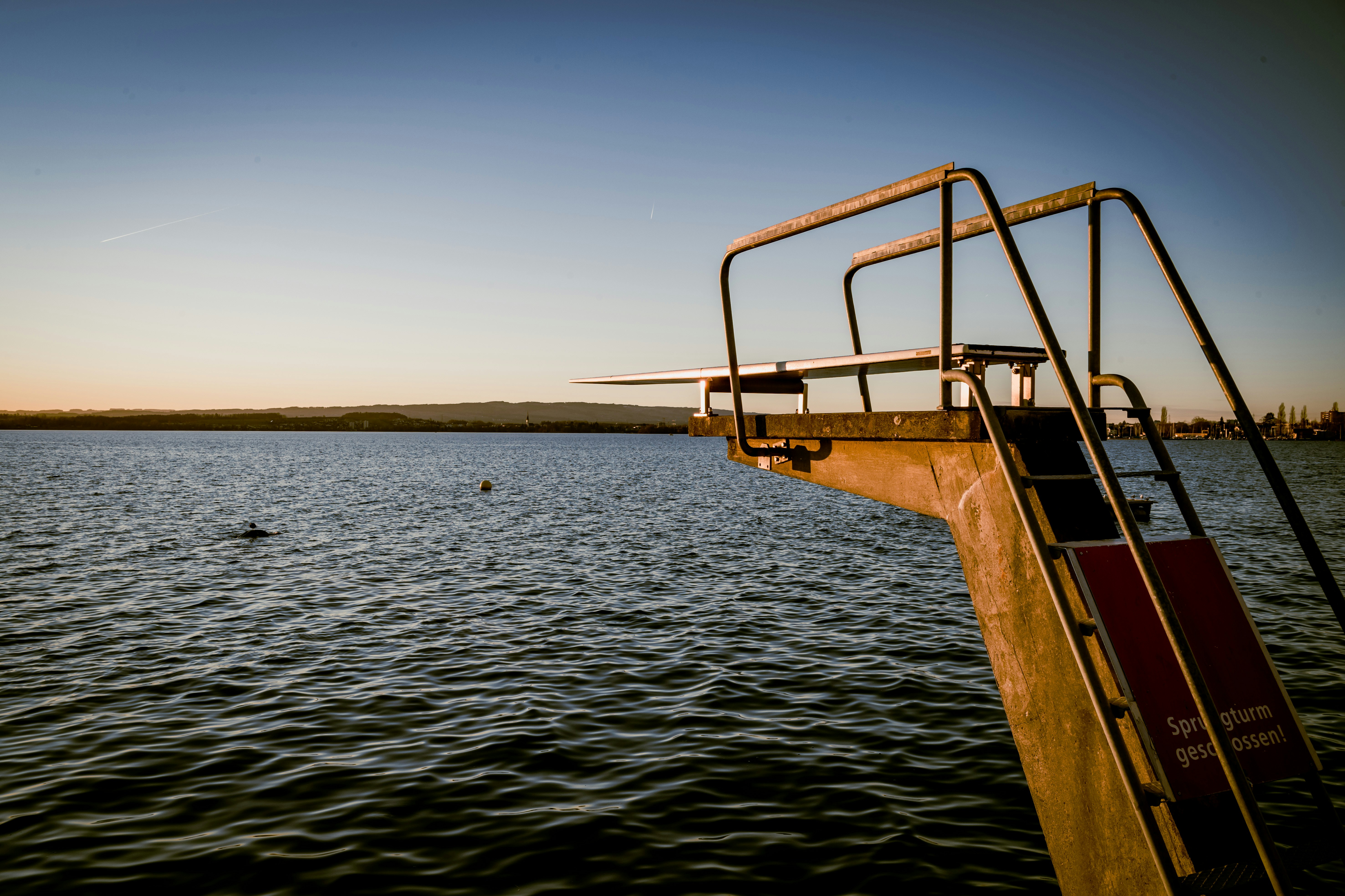 Diving board silhouetted against tranquil lake at sunset.