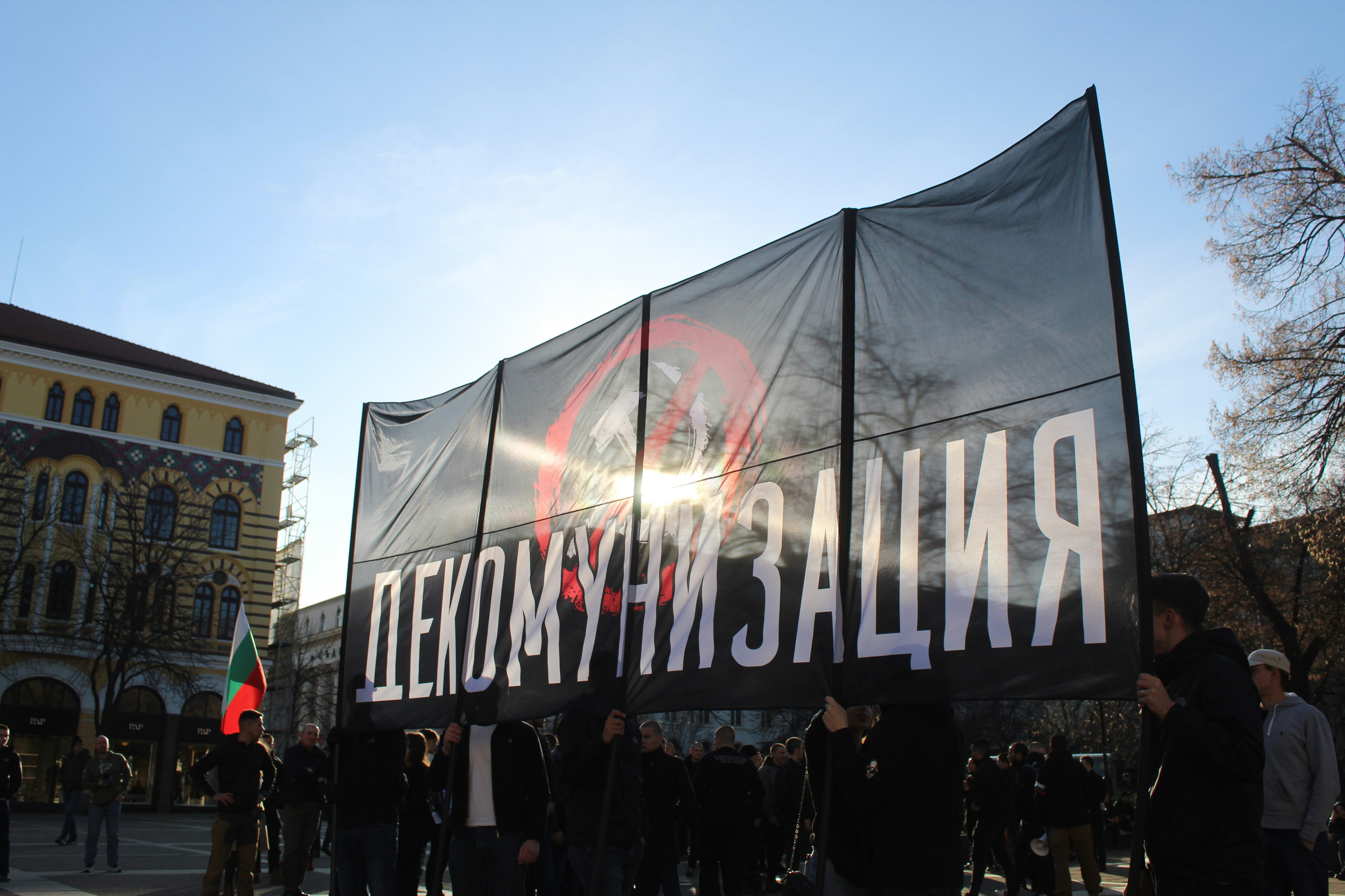 Protesters gather with a large banner against the backdrop of the sun, highlighting a call for decommunization.