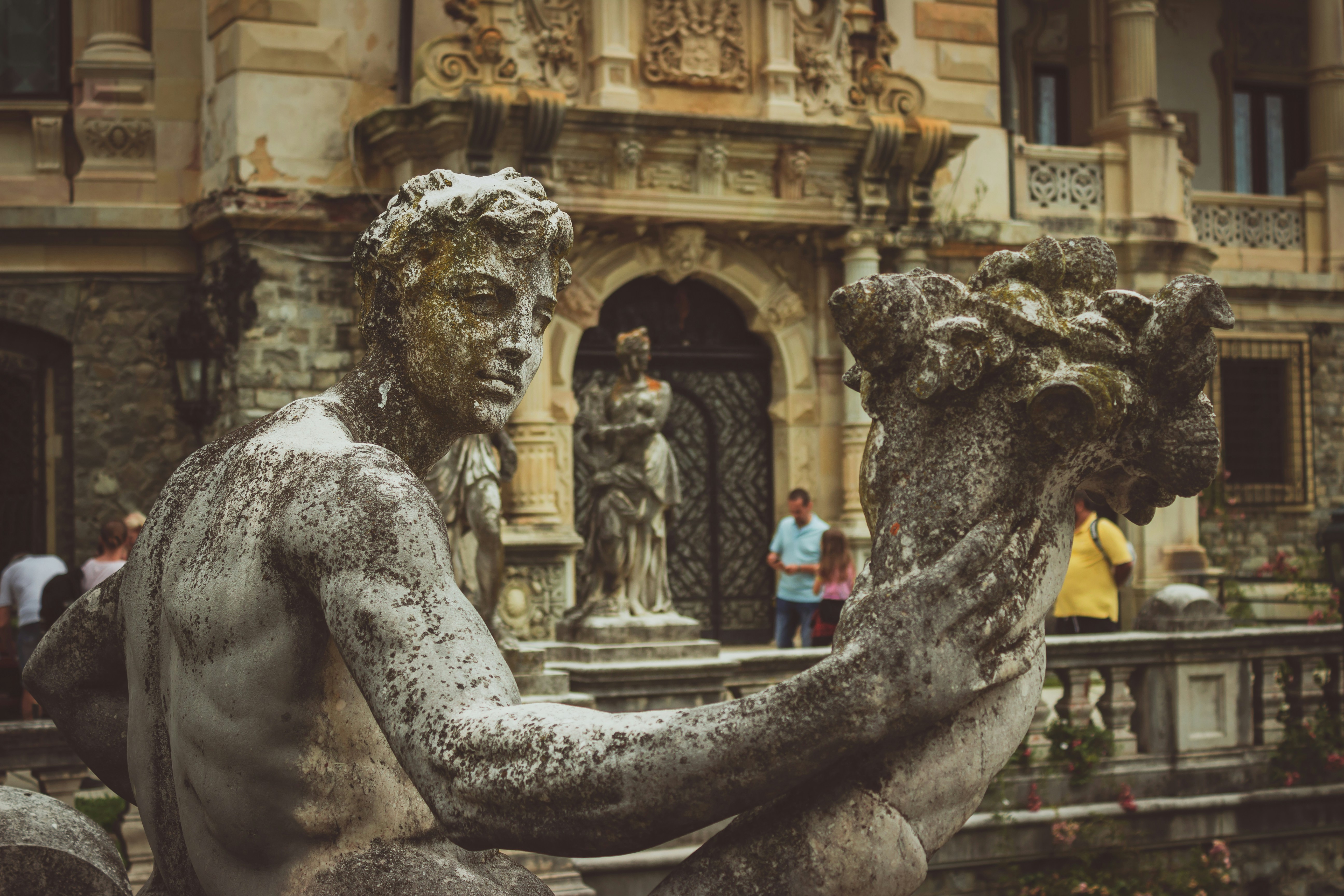 Weathered stone statue gazing across a grand historical building facade, with people in the background.
