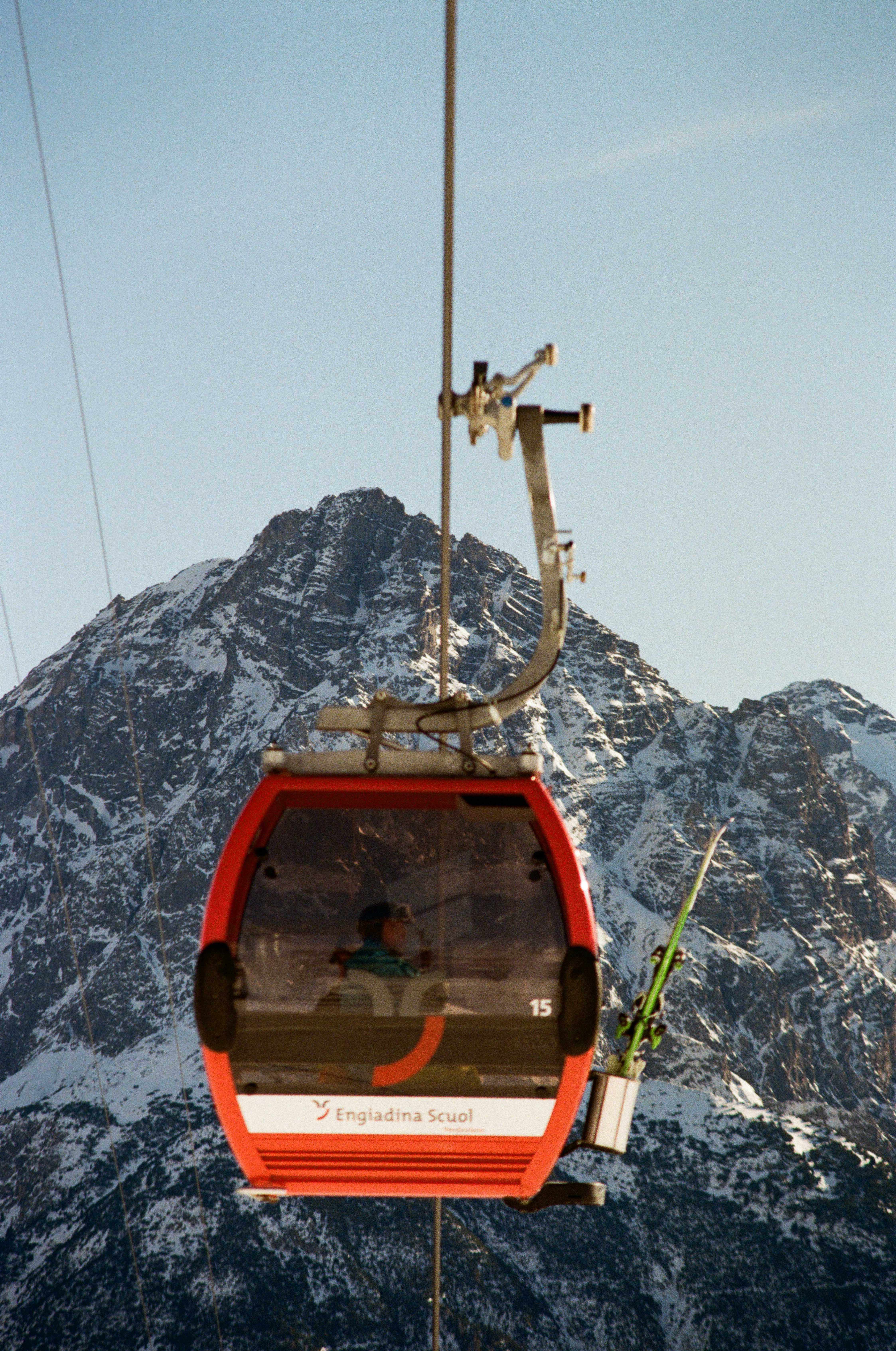 Red gondola cabin suspended by a cable glides above snow-dusted Alpine peaks. Passengers appear through the cabin's glass, set against a pale blue sky.