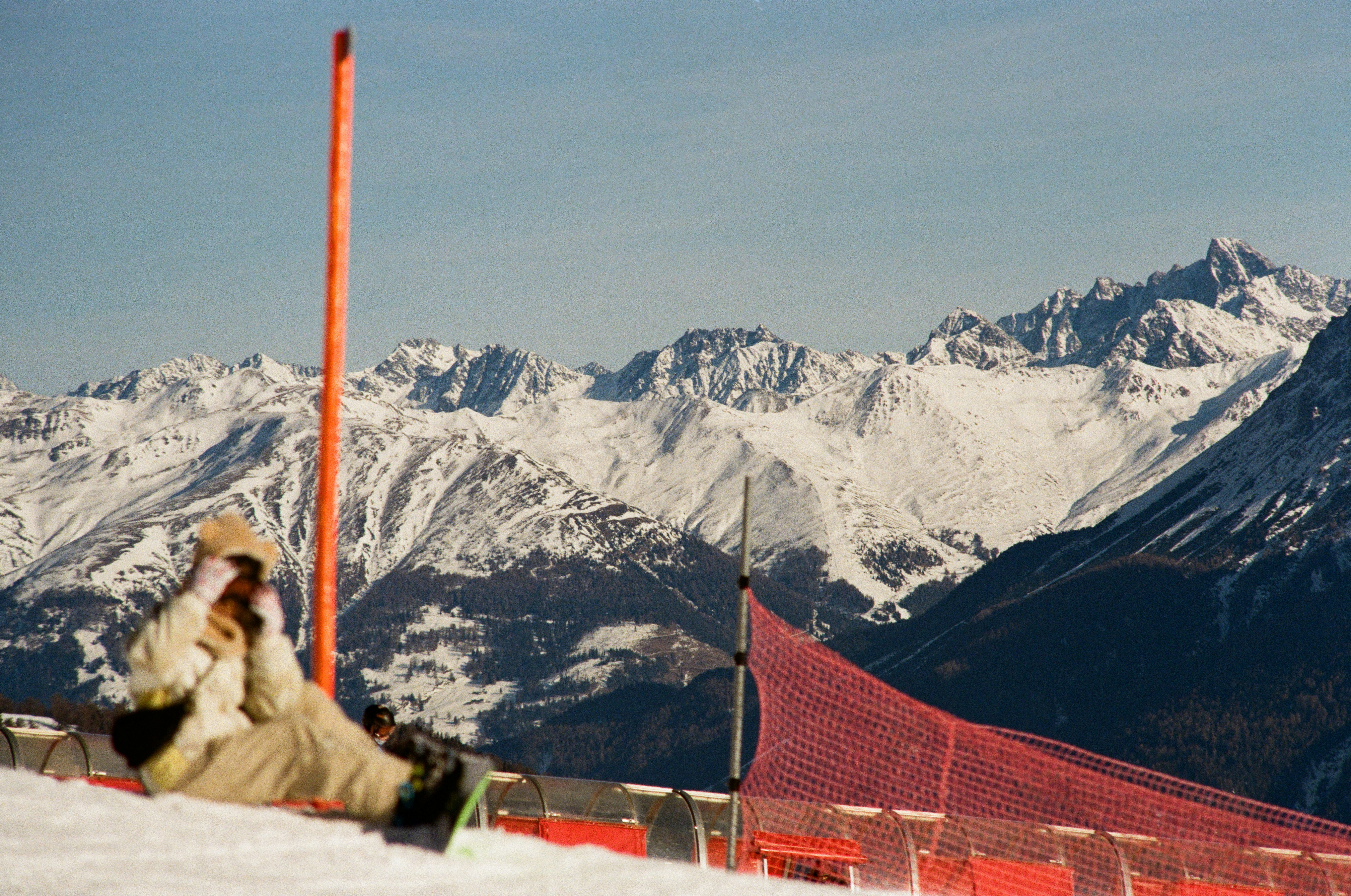 Snowboarders resting on a snowy slope with the Alps and red safety netting under a clear blue sky.