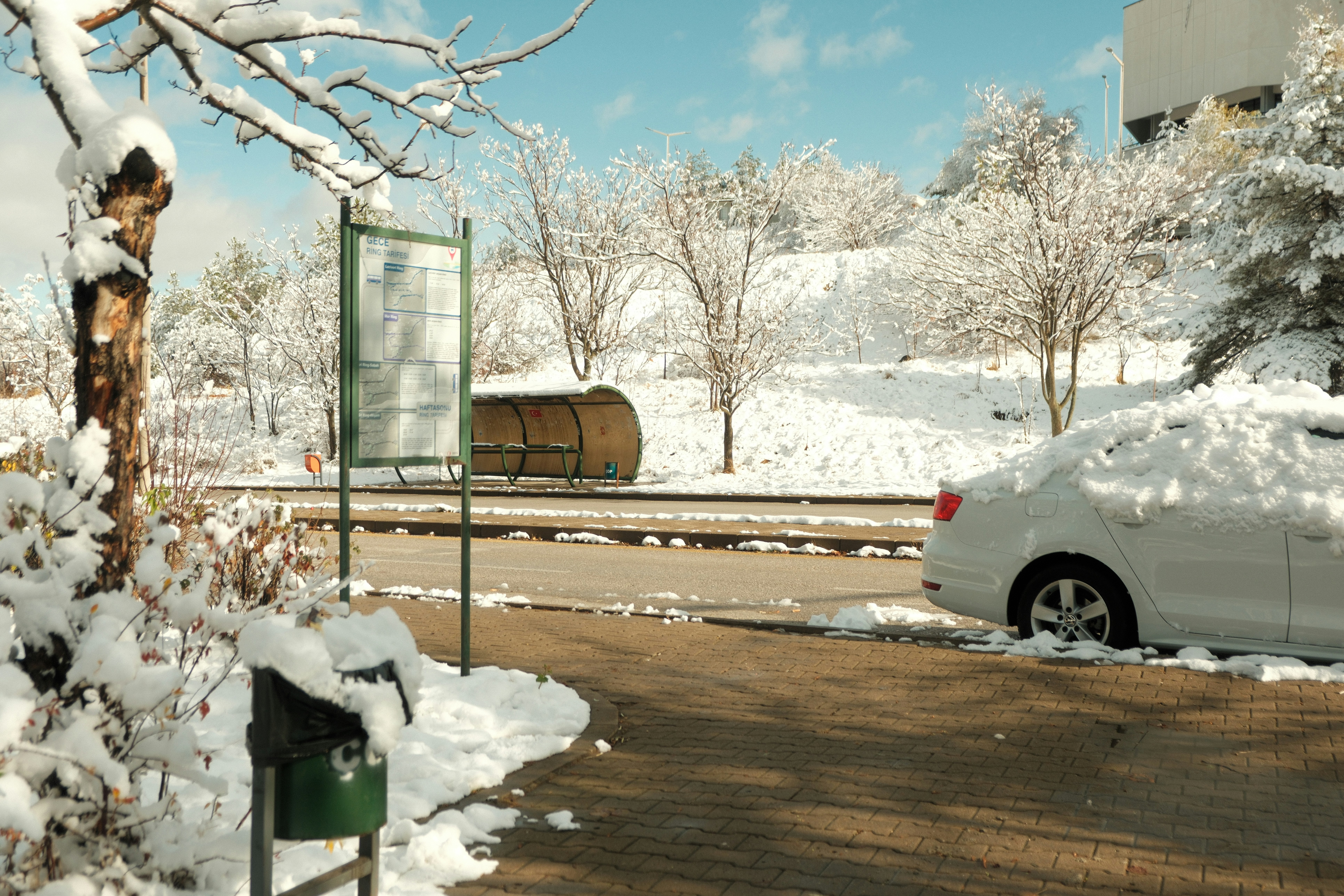 Snow-covered bus stop with parked car and bare trees under a clear blue sky.