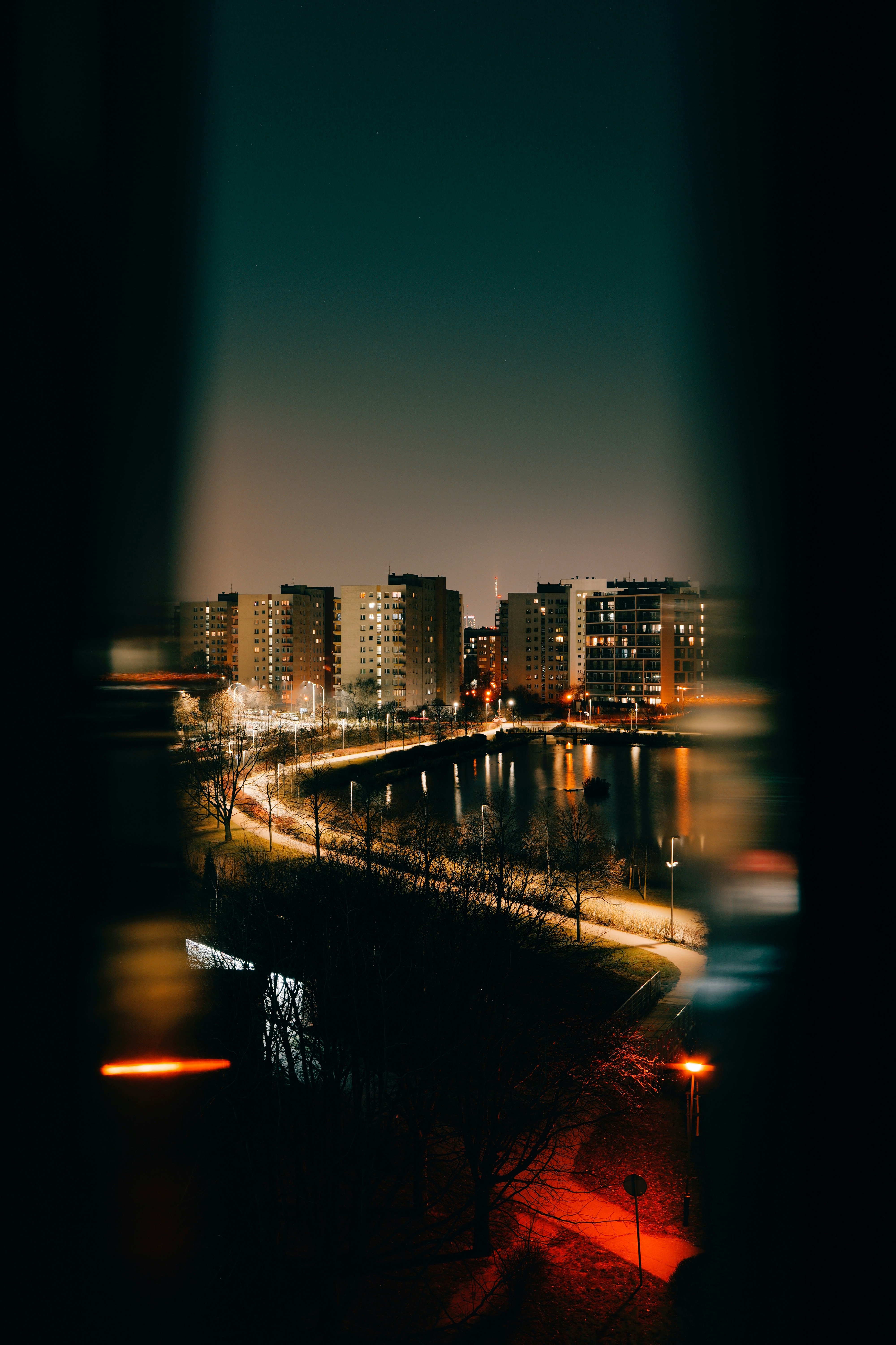 Nighttime cityscape photographed through a narrow window frame, using a long exposure to blur traffic and render warm reflections along the river.