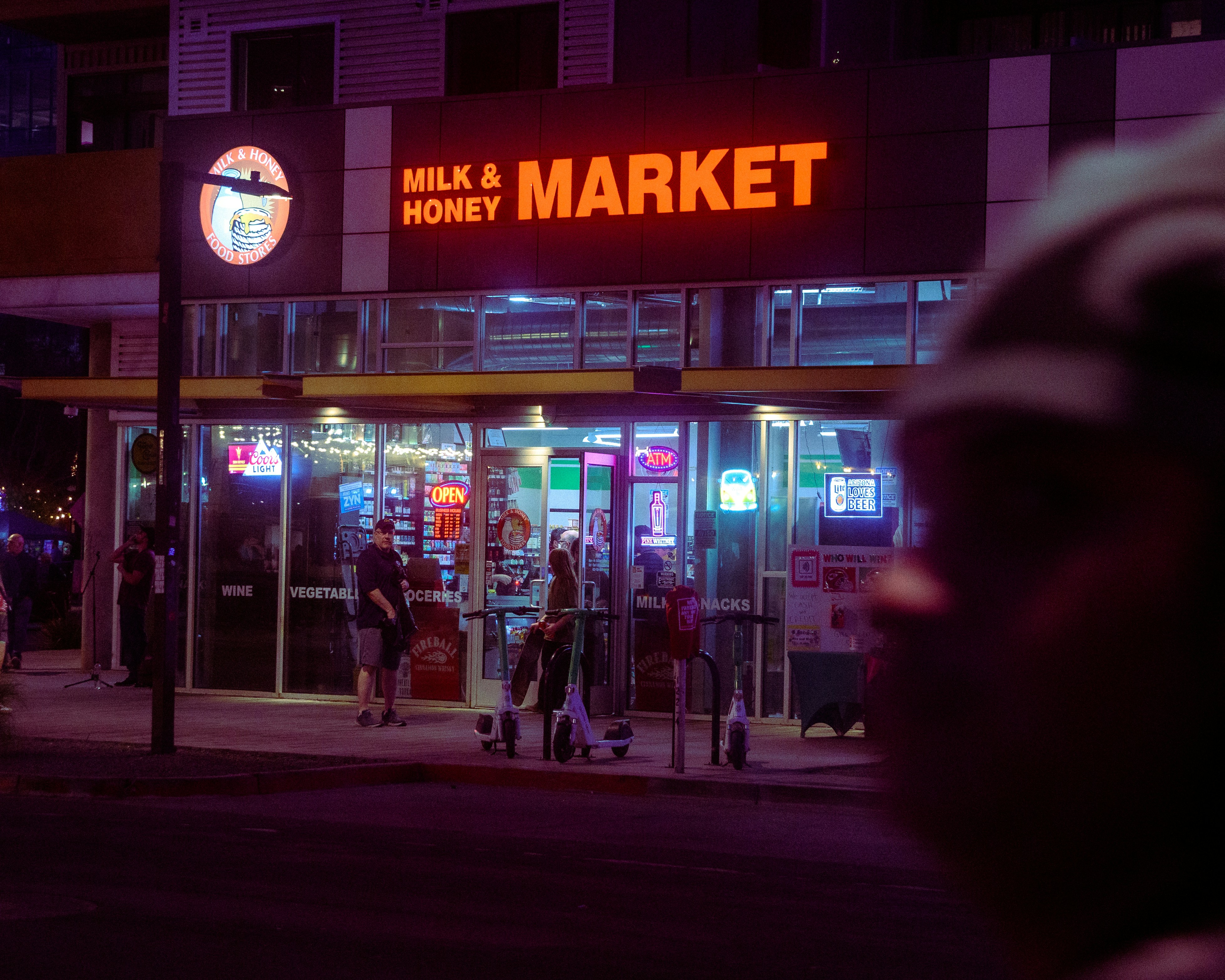 Neon Night Market: Glass storefronts and a blurred silhouette at the corner