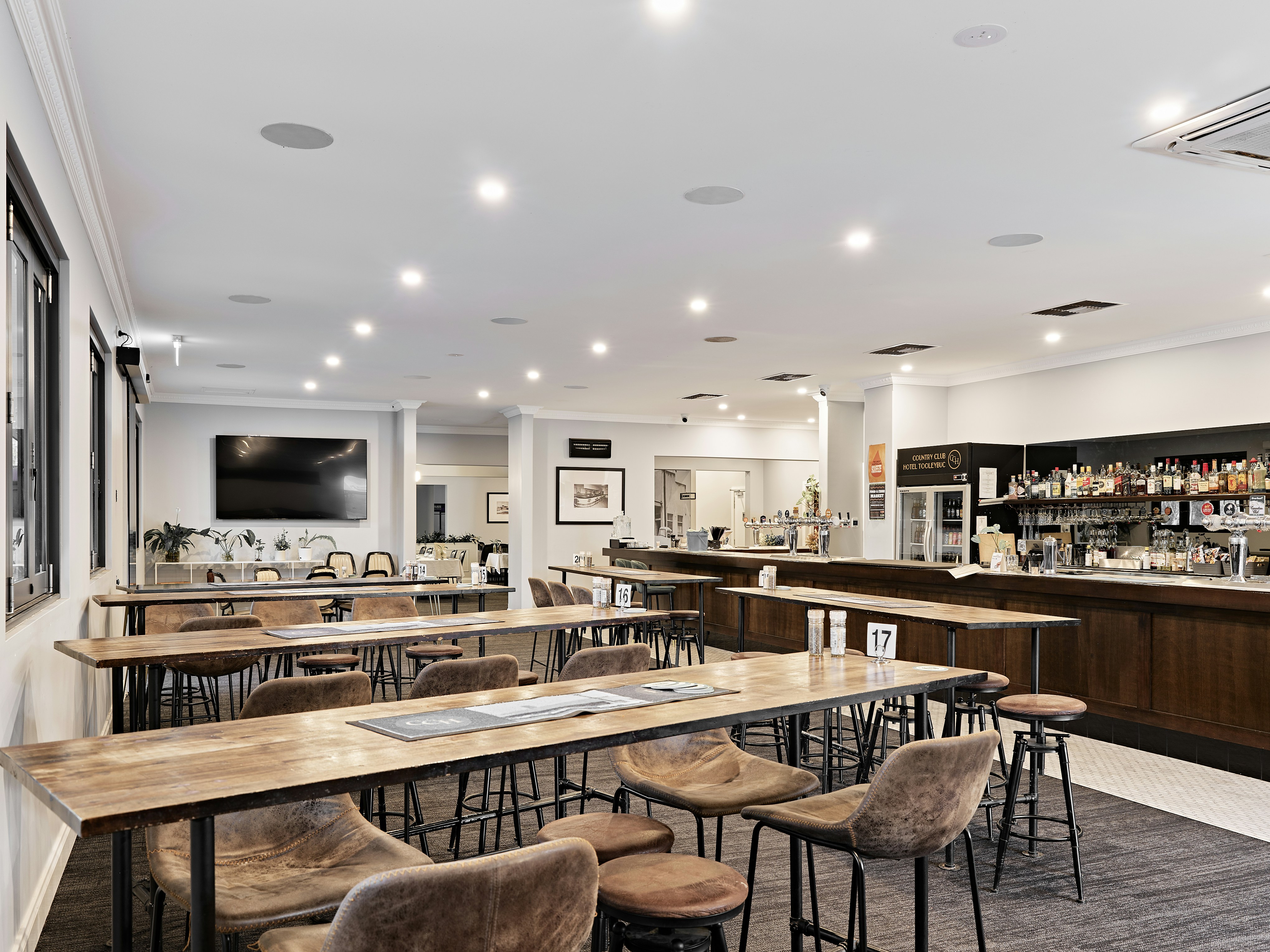 Modern bar interior with long wooden tables and plush brown chairs under soft recessed lighting, leading to a well-stocked bar.