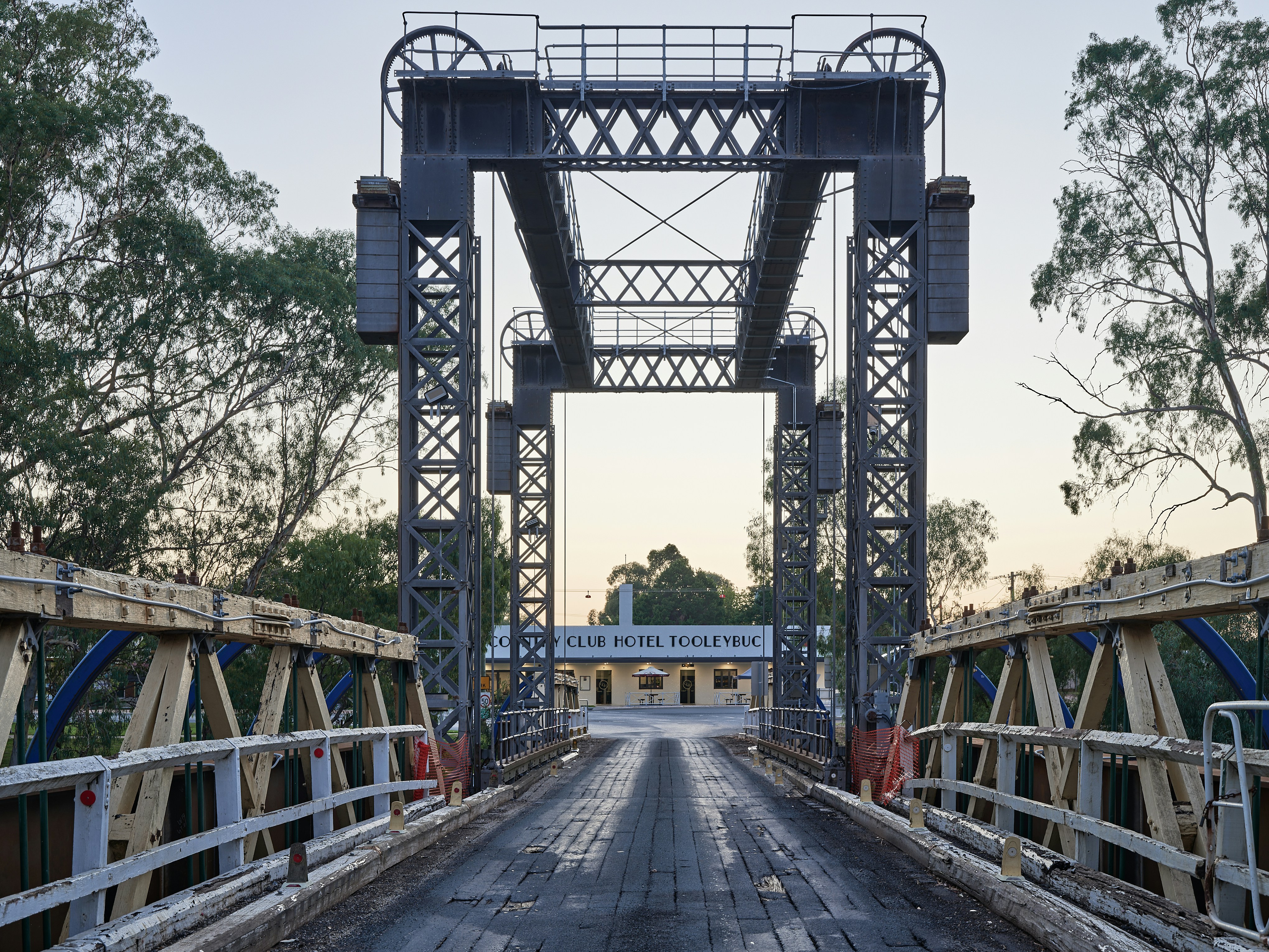 Symmetrical iron bridge framed by lush trees, with cool steel tones contrasting against the warm evening sky.