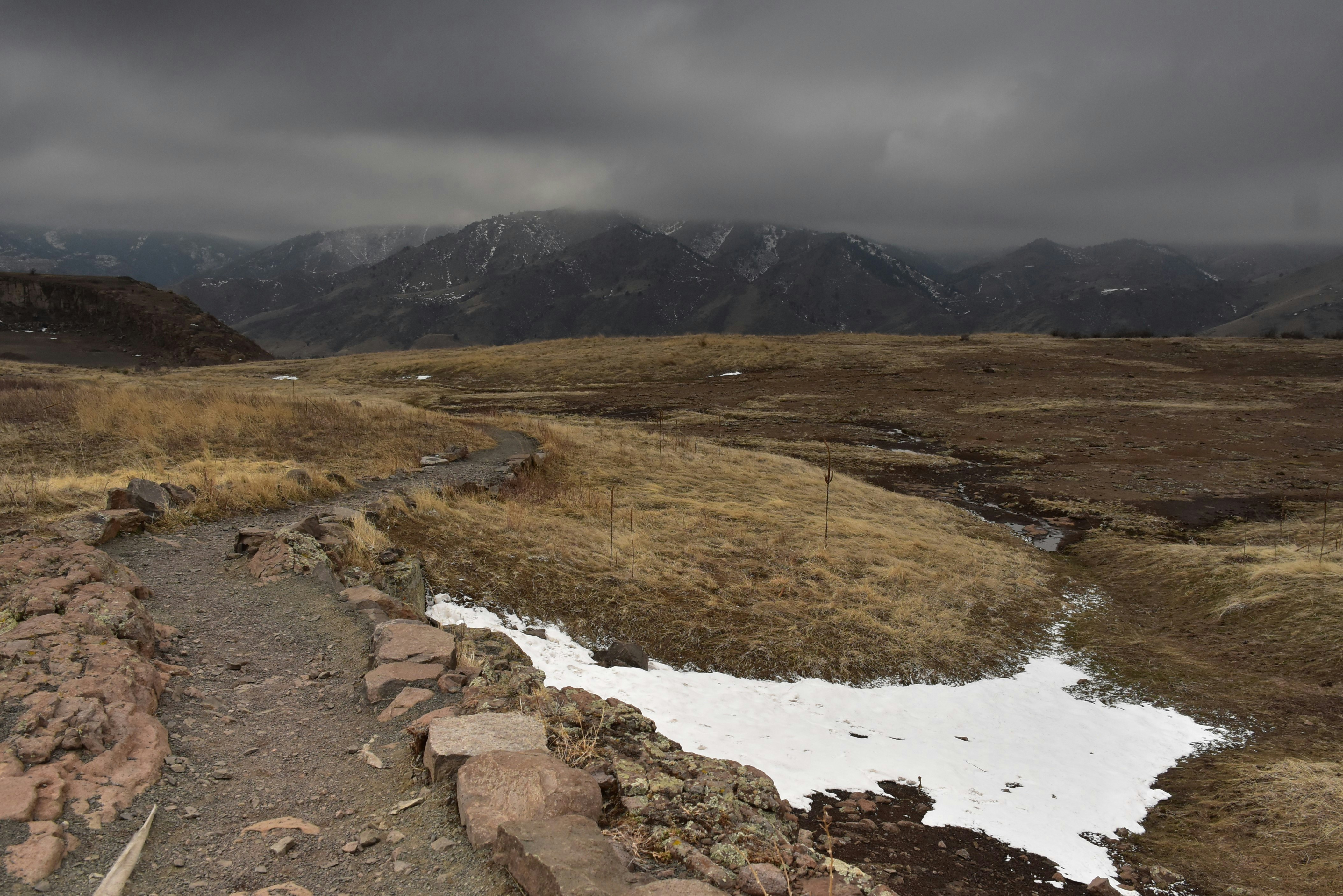 Winding dirt path through a grassy field beneath overcast mountains.