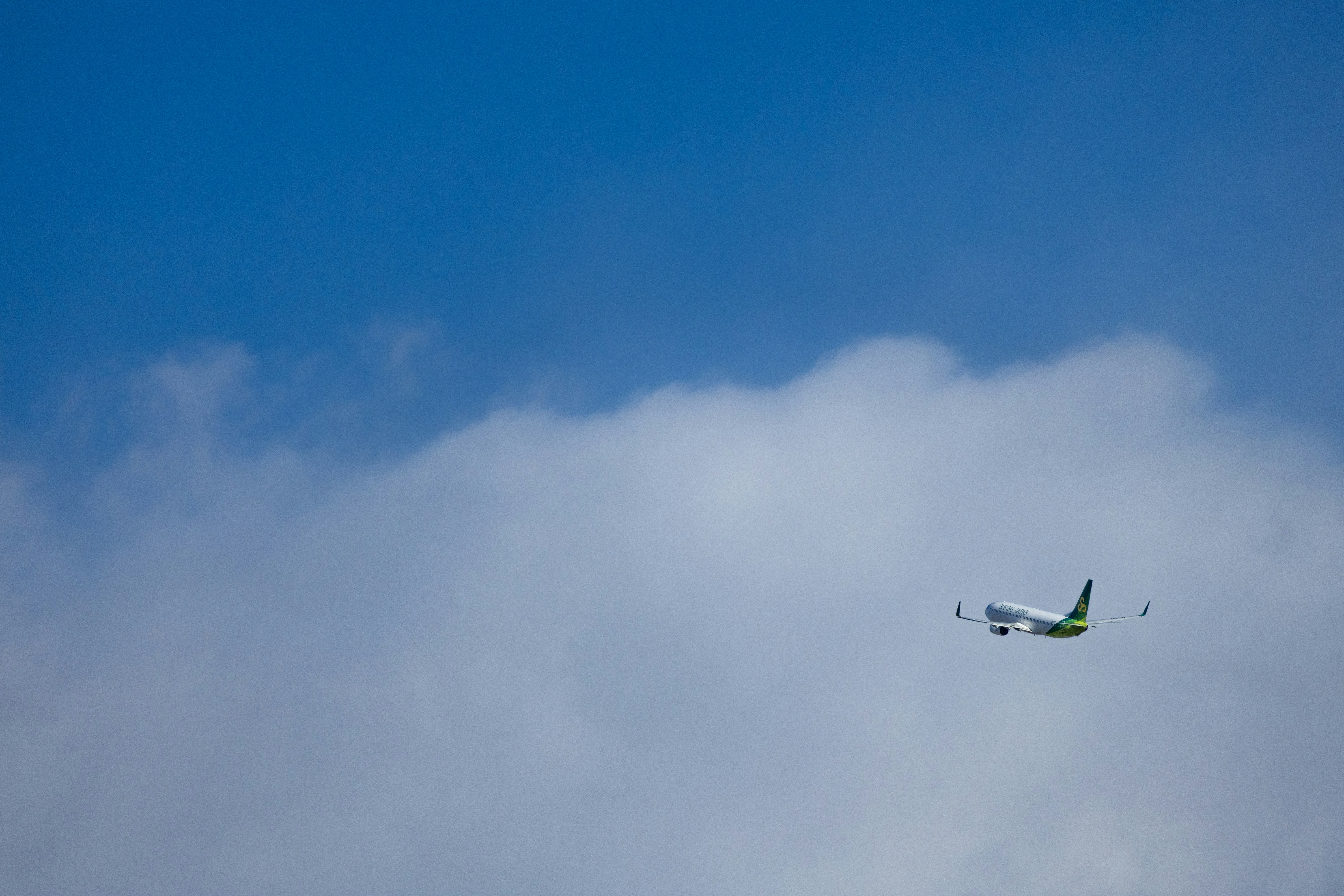 Airplane flying through a blue sky with soft white clouds, a green tail accent contrasting the aircraft's white body.