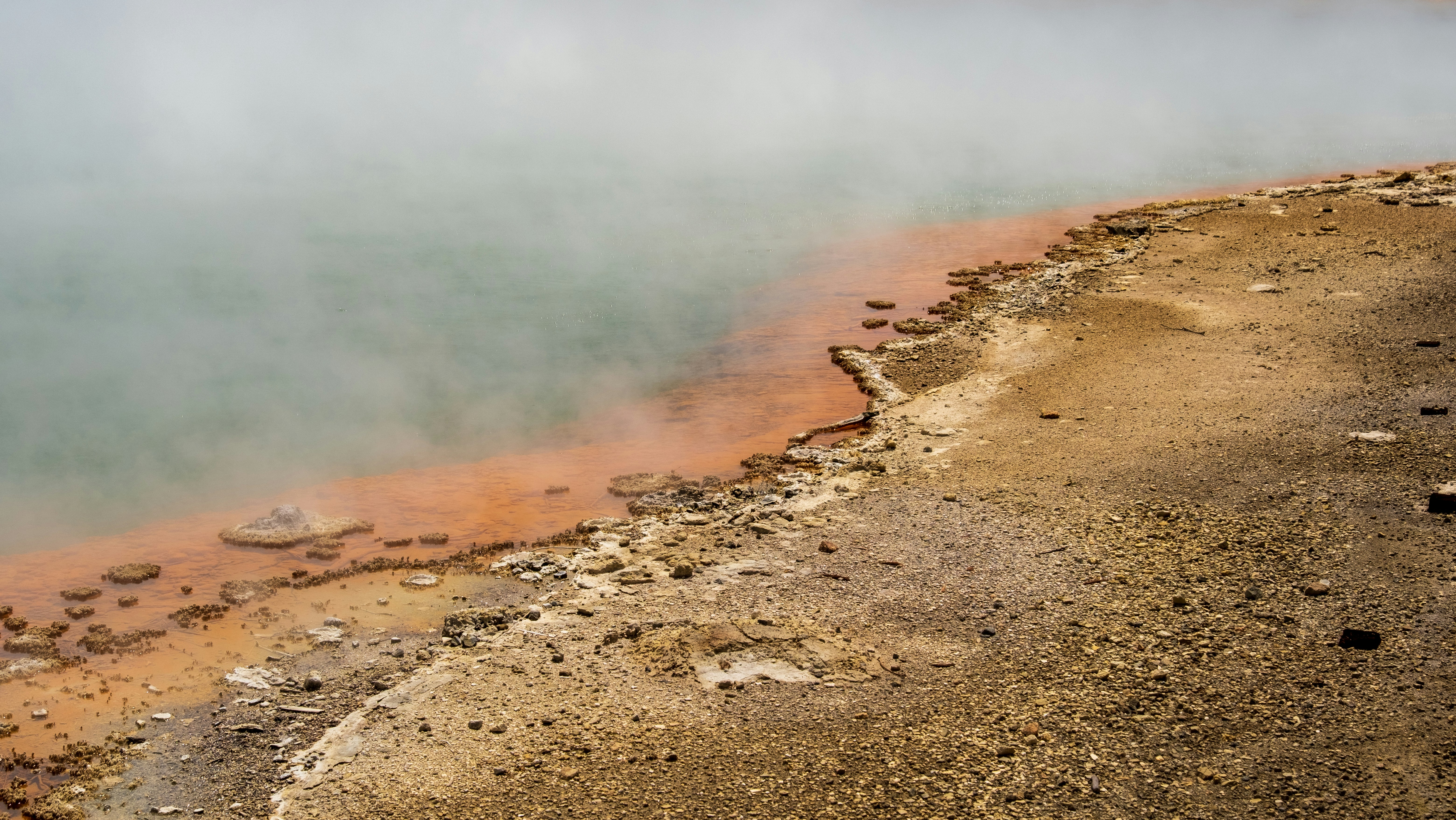 Geothermal pool's vibrant orange shoreline meets a veil of mist, contrasting textures and tones.