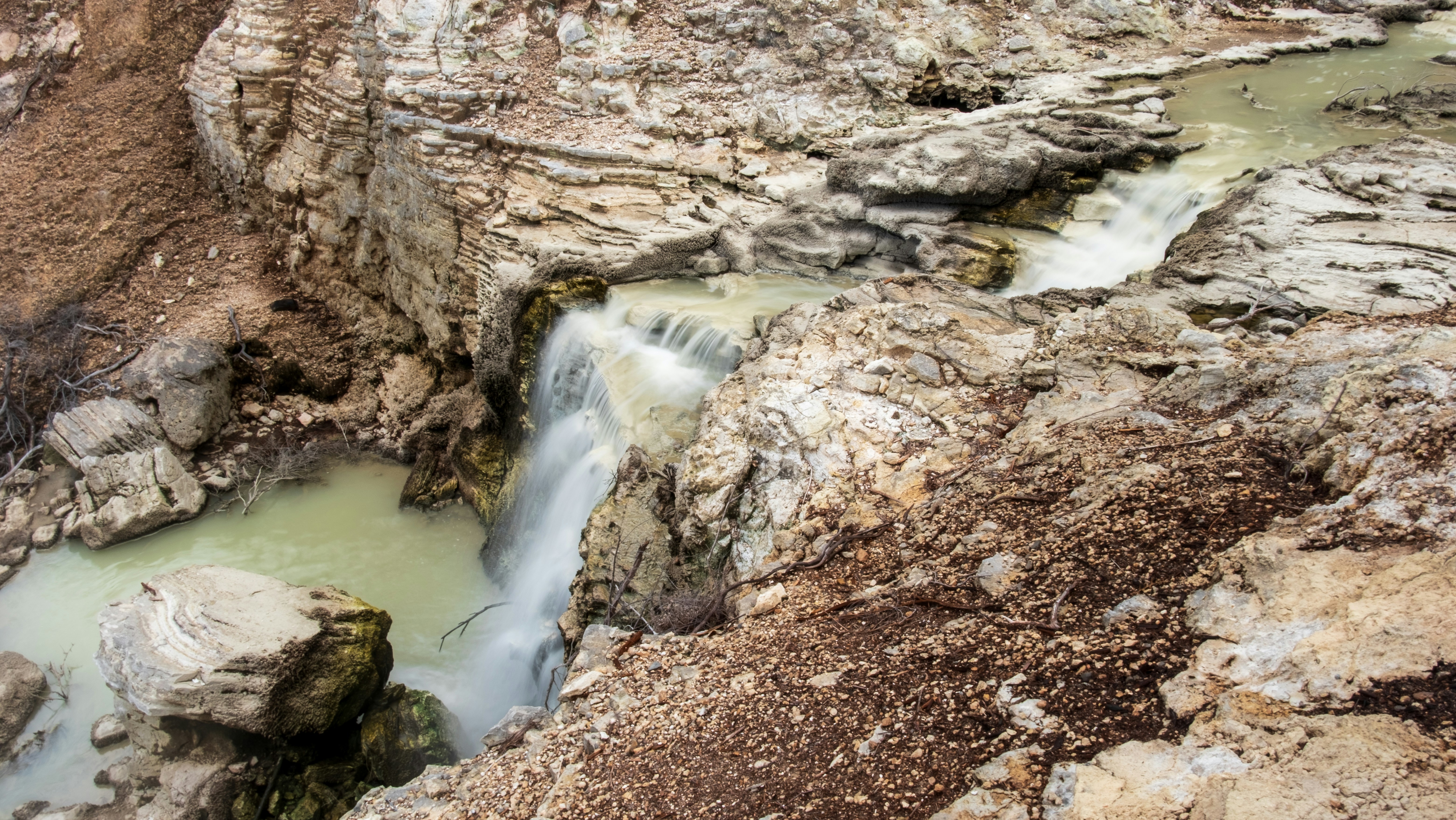 Waterfall gently flowing over weathered rocks with earthy tones and milky green water.