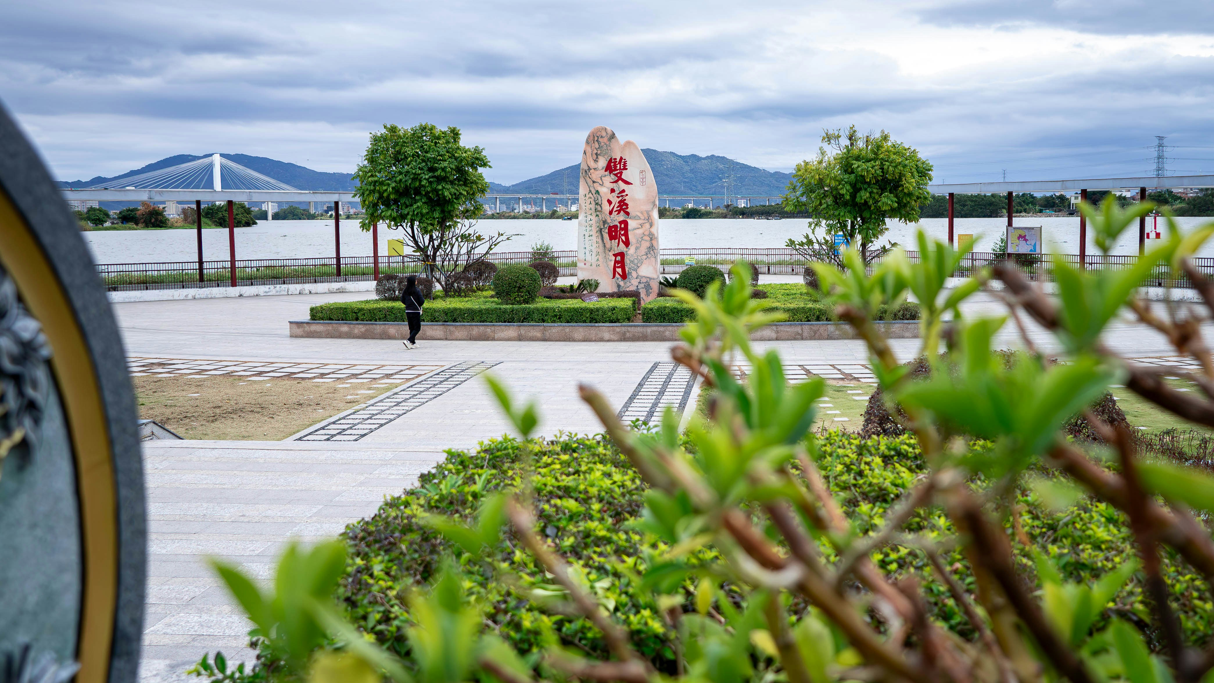 Stone monument with red inscriptions stands amid lush greenery near a serene river, framed by distant mountains under a cloudy sky.