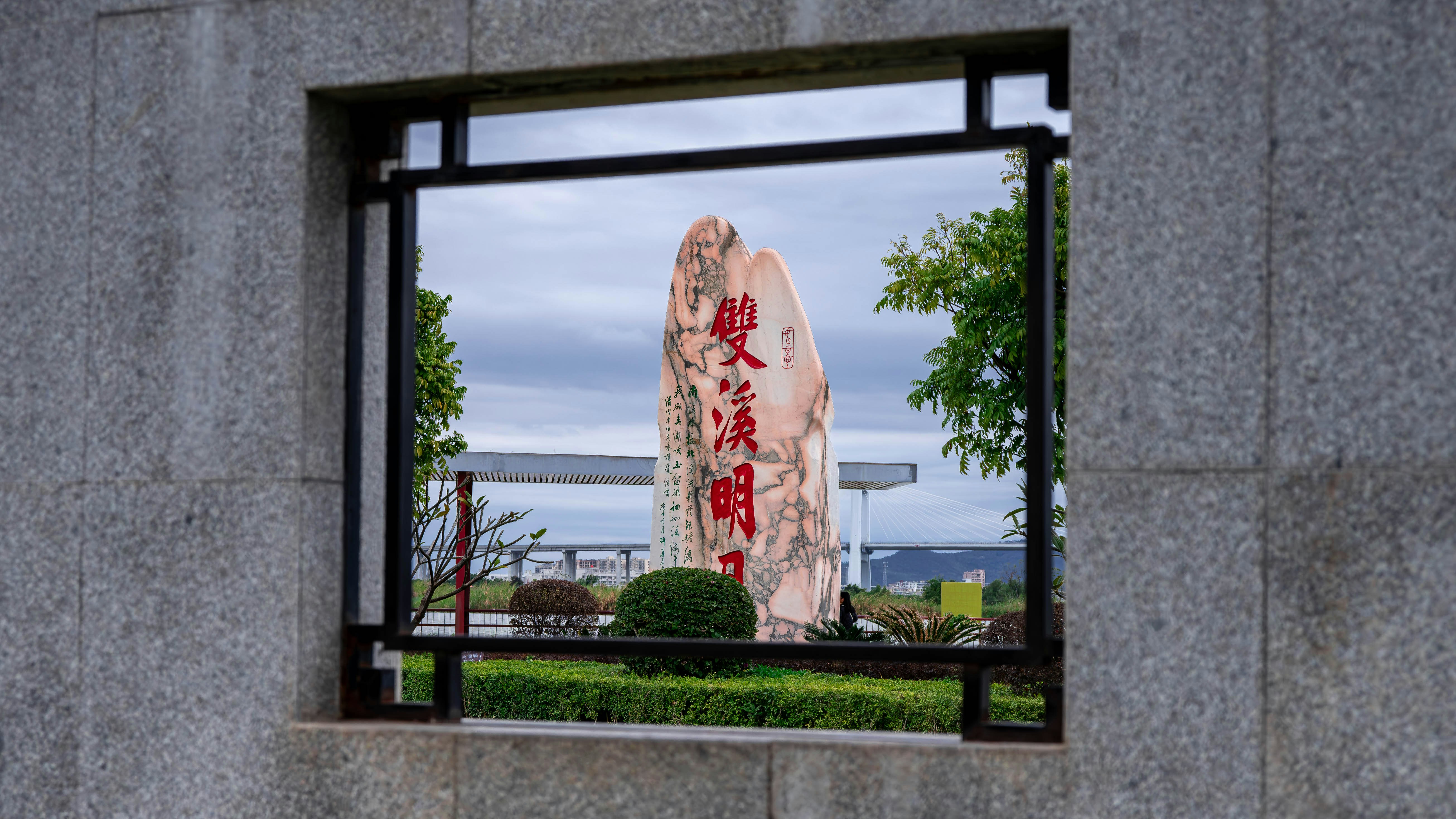 Stone monument framed by an architectural window under overcast skies, surrounded by greenery.