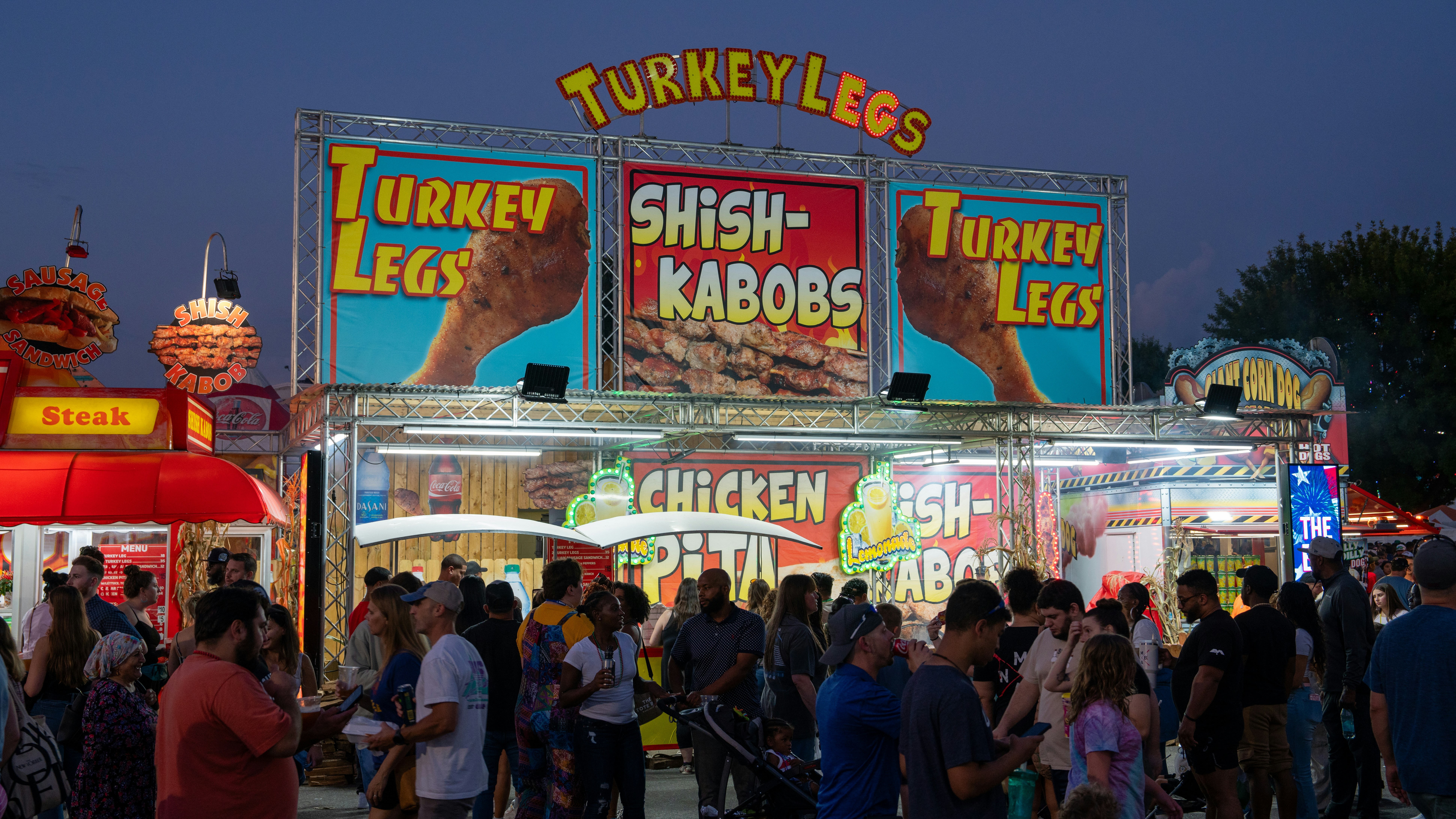 Busy fairground food stand with bright signs for turkey legs and shish-kabobs against a dusky sky.