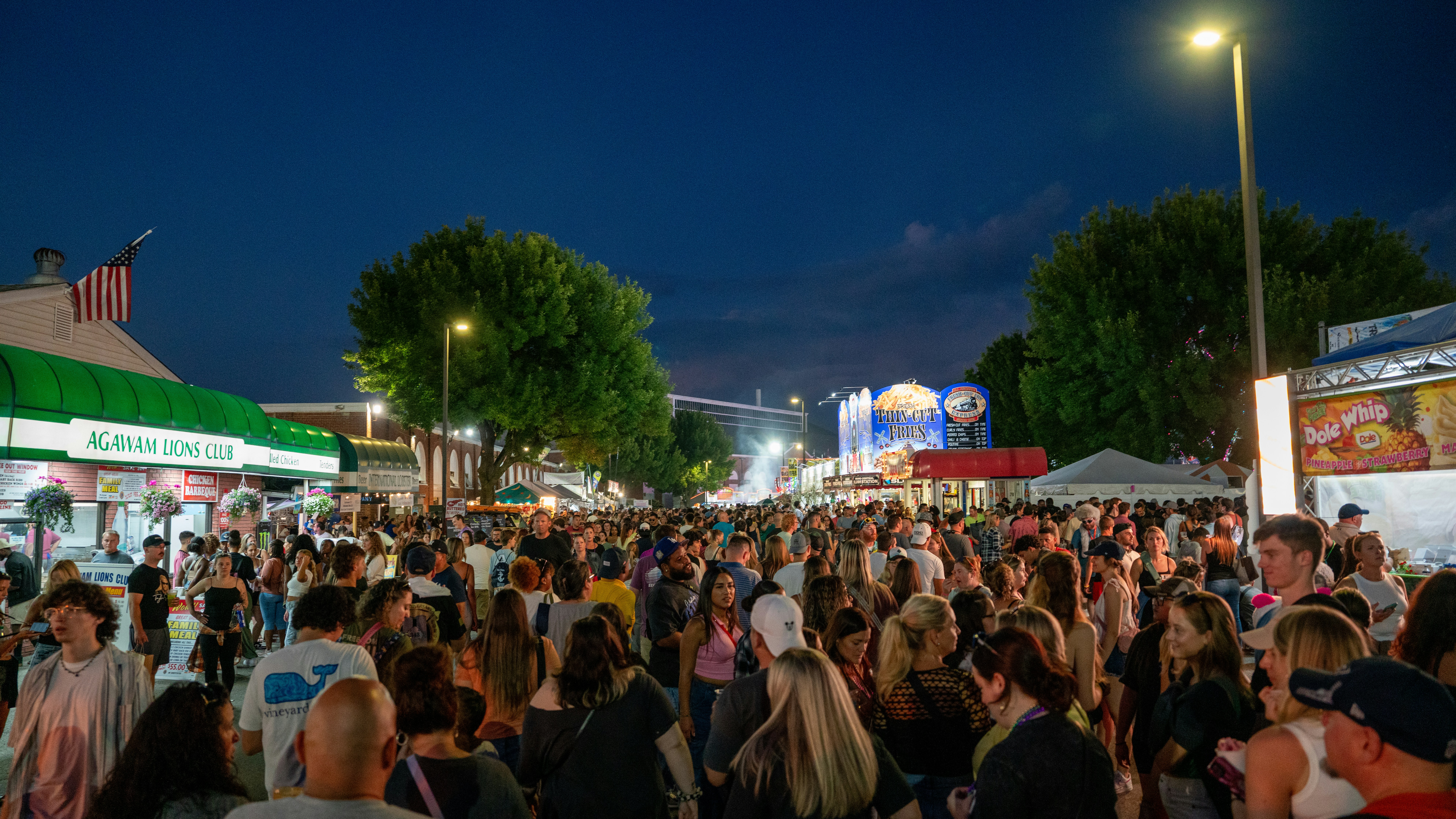 Crowd enjoying a lively summer fair under twilight skies, illuminated by colorful booth lights and streetlights.