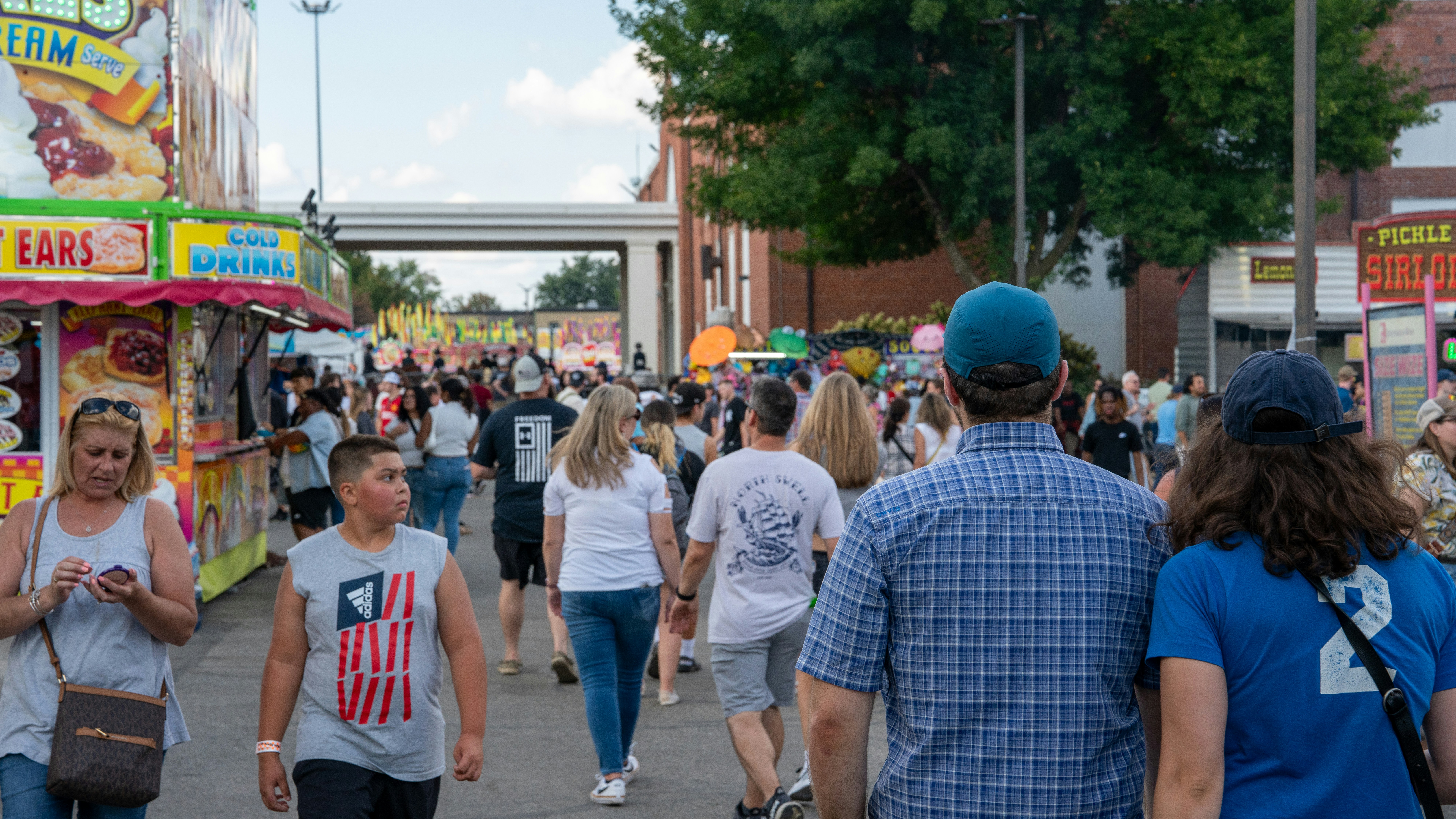 Crowd enjoying a sunny day at a bustling fair with colorful food stalls and vibrant signage under a soft, natural light.
