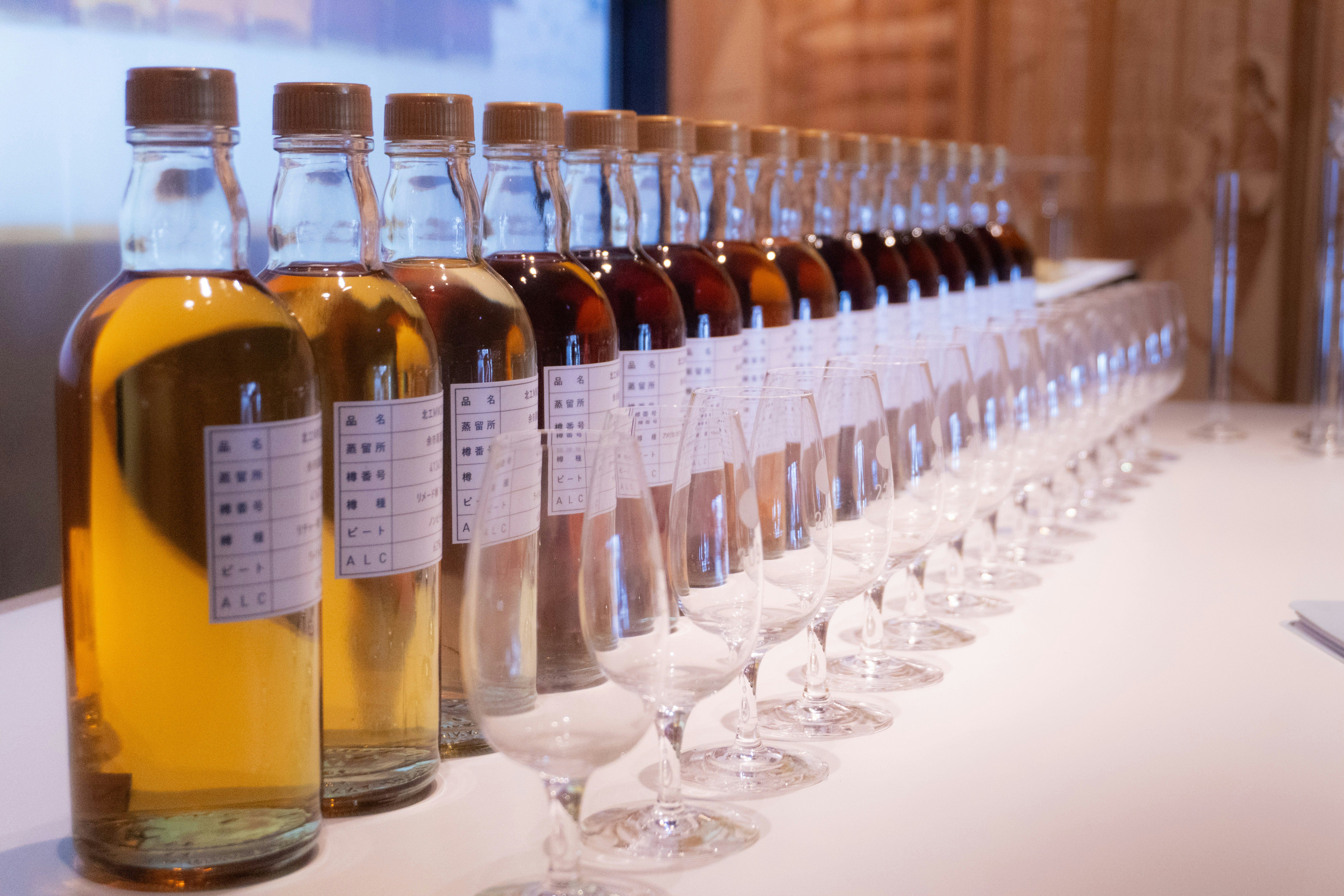 Row of bottles with amber and golden liquids aligned with empty glasses under warm lighting.