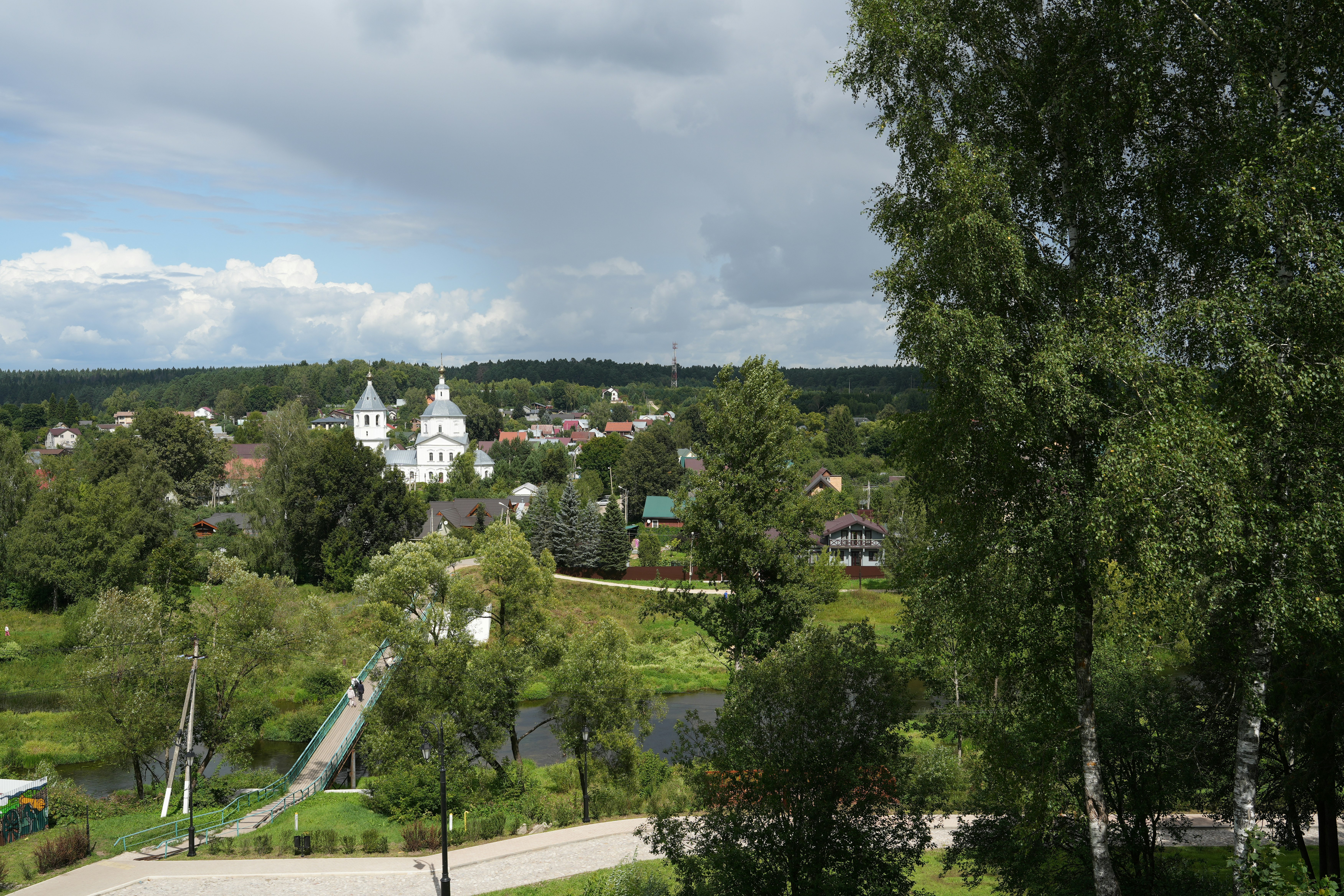 Scenic view of a church amidst lush greenery with a river and pathway under a partly cloudy sky.