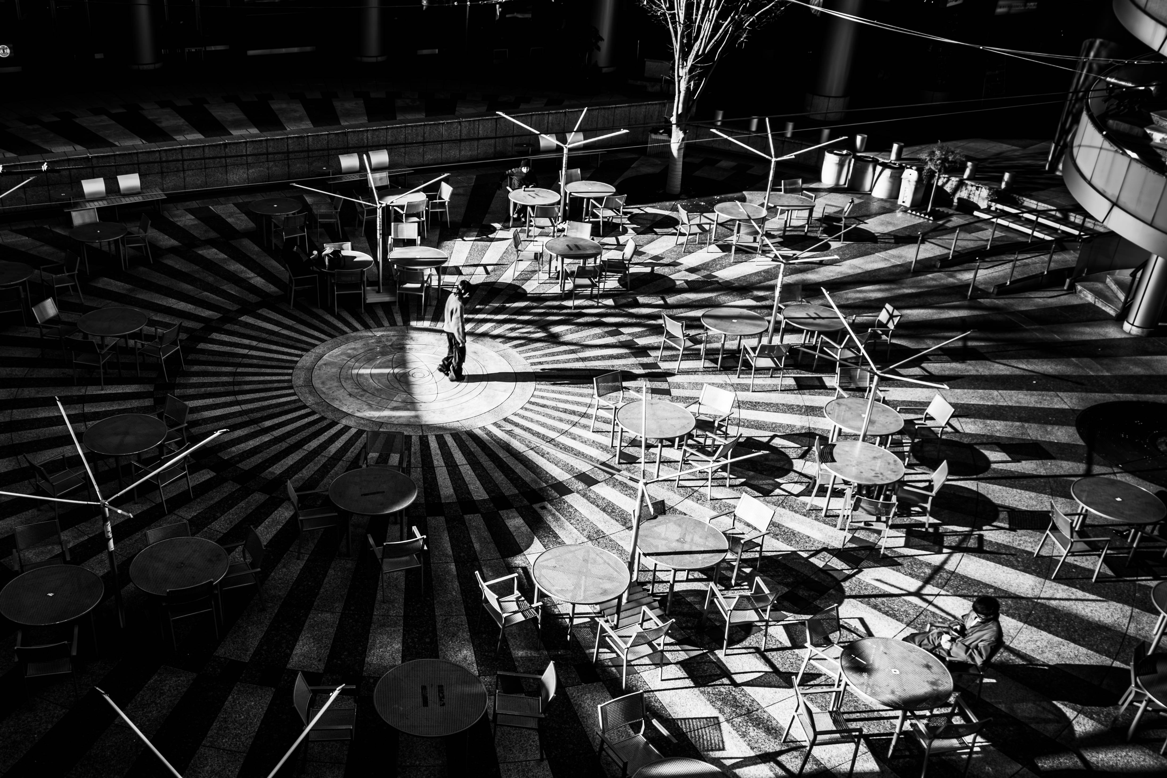 Black and white photo of a lone figure in a plaza with a radial pattern of light and shadows, surrounded by empty tables and chairs.