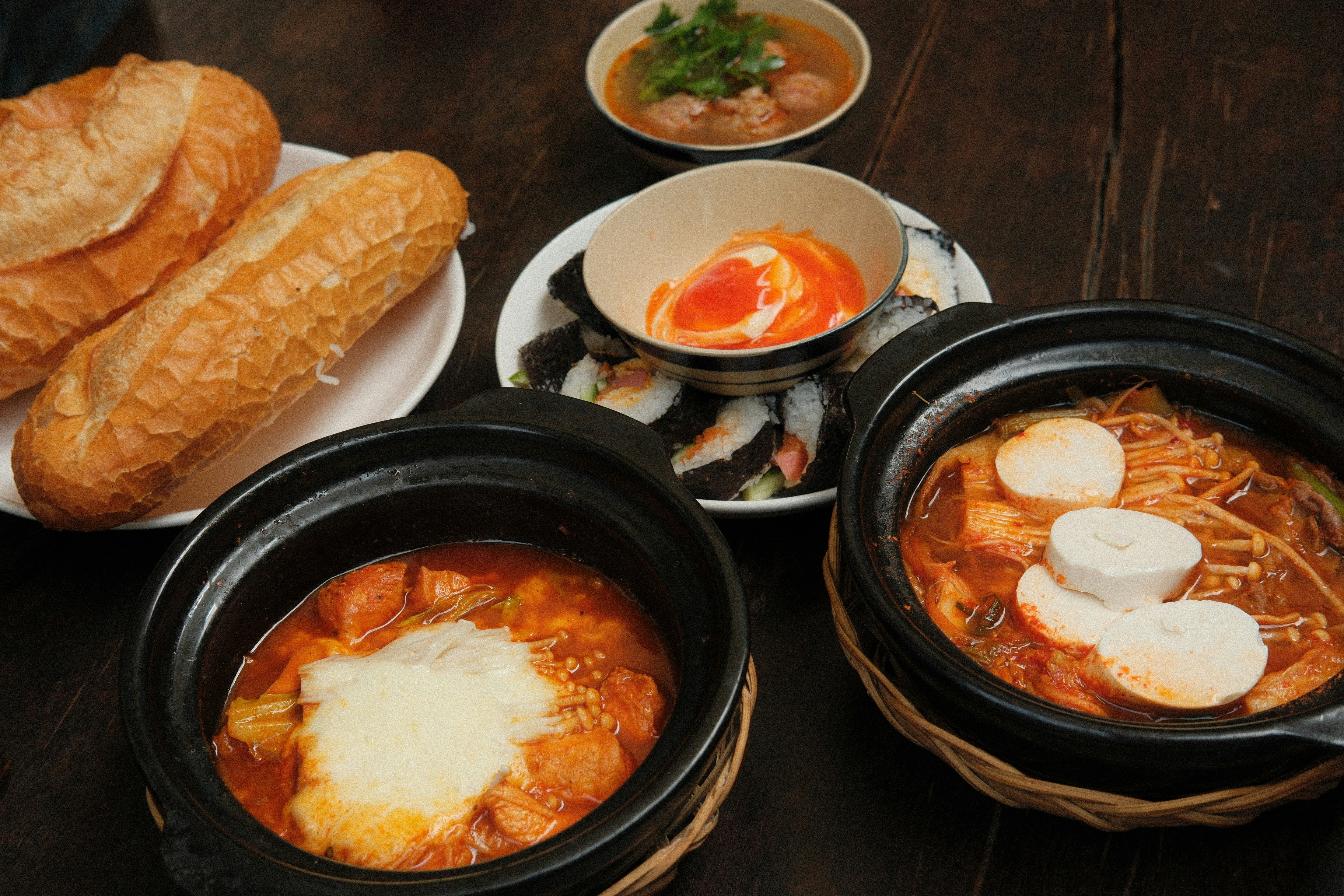 Steaming Korean stews with tofu and vegetables next to baguettes and sushi rolls topped with a runny egg on a wooden table.