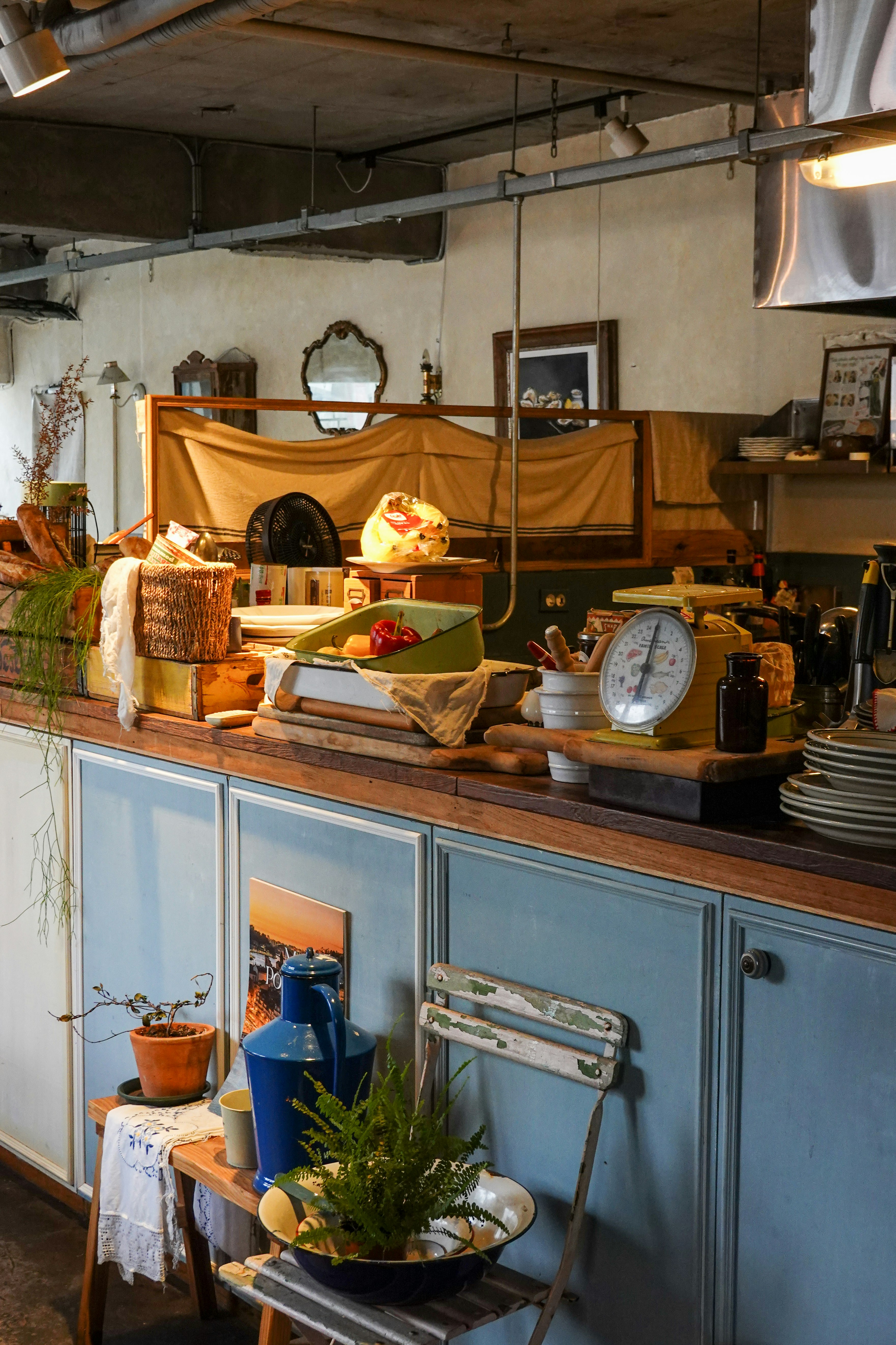 Cozy rustic kitchen counter cluttered with baskets, fresh produce, and a vintage scale atop blue cabinetry. A cafe-style prep area bathed in warm ambient light.