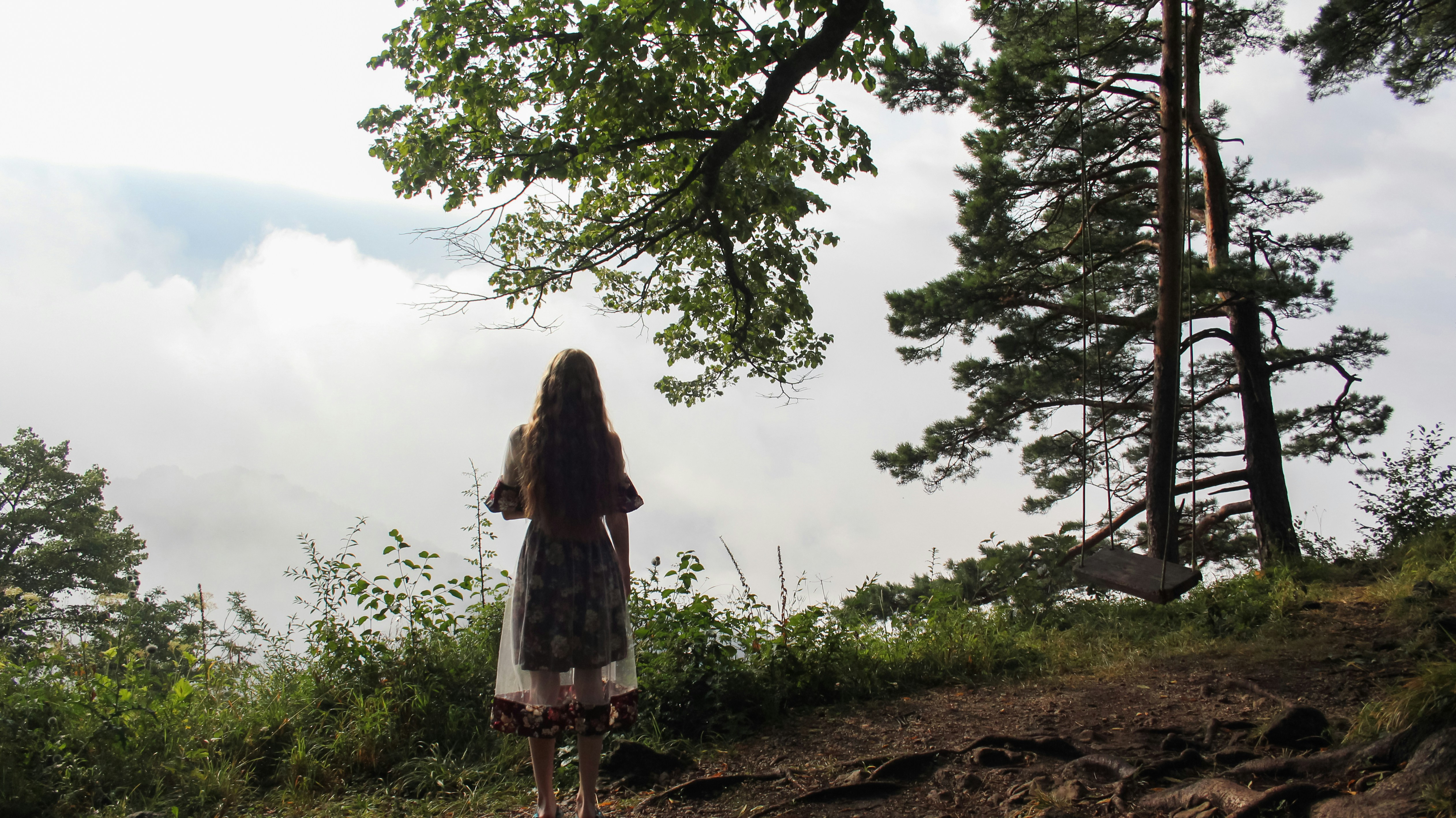 Girl stands at forest edge amidst mist and lush greenery, framed by trees and a swing. Soft light enhances the tranquil scene.