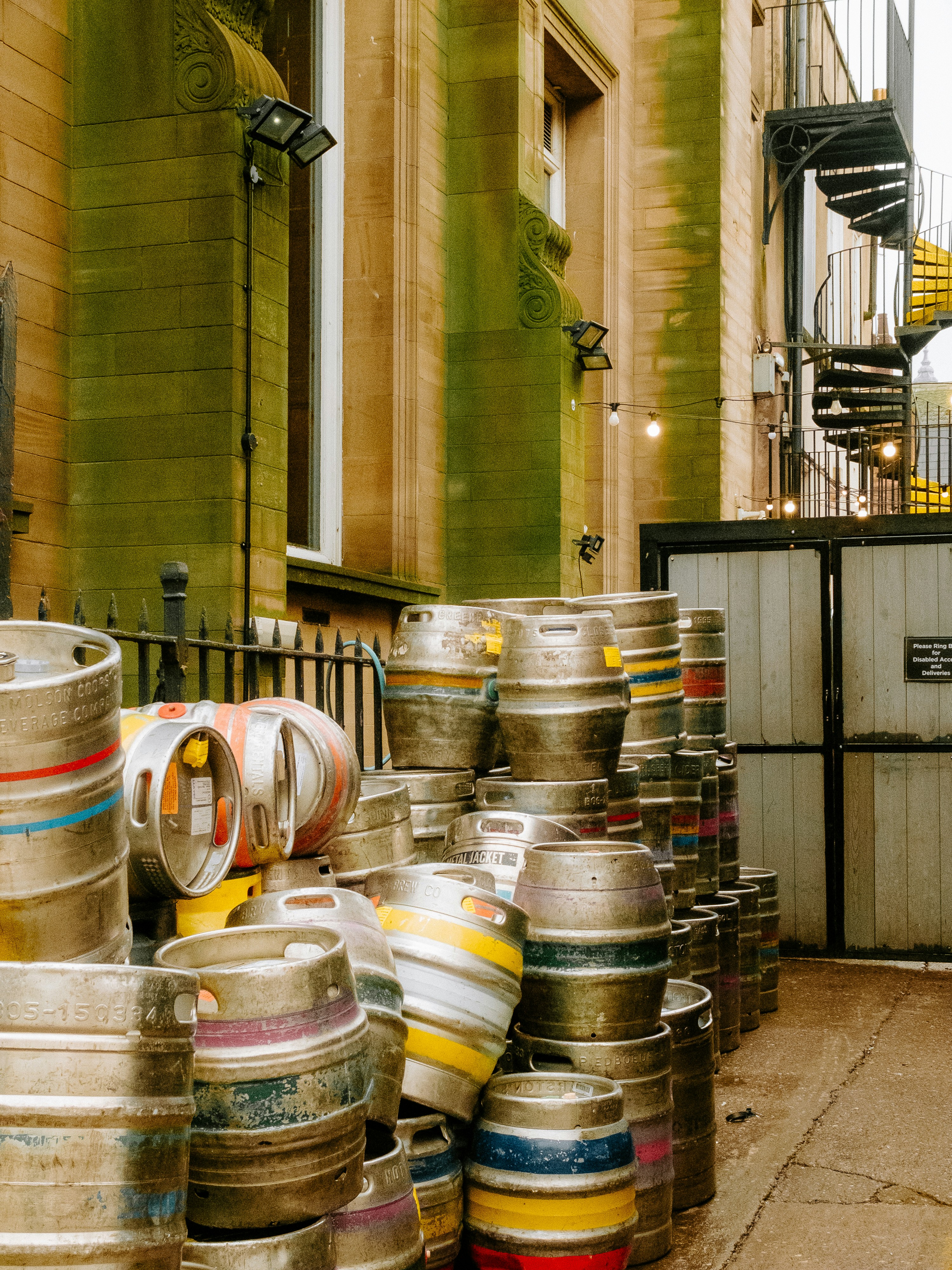 Colorful beer kegs stacked against a textured stone wall, with a spiral staircase and warm lighting in the background.
