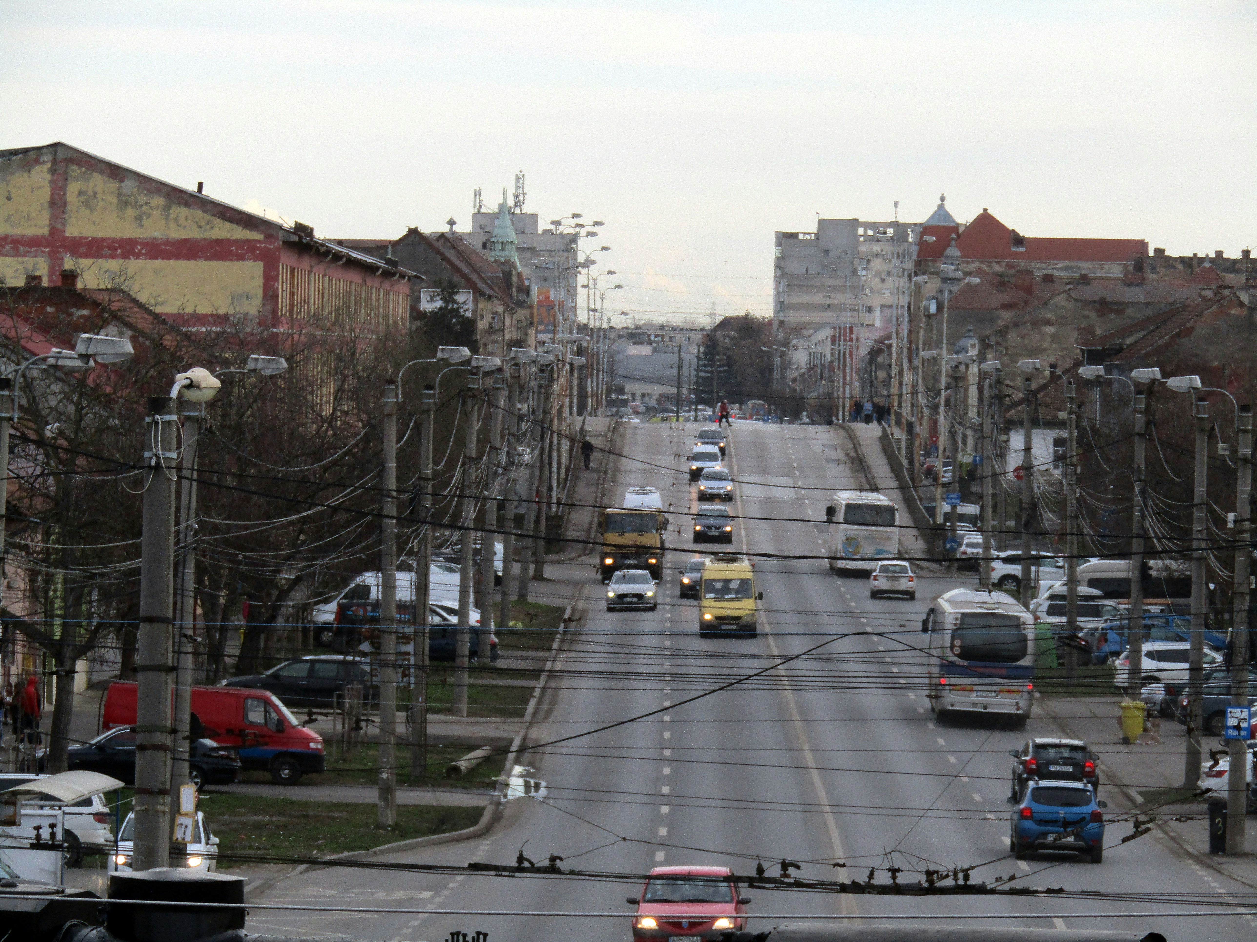 Busy city street with cars ascending a hill, flanked by aged buildings and power lines under an overcast sky.