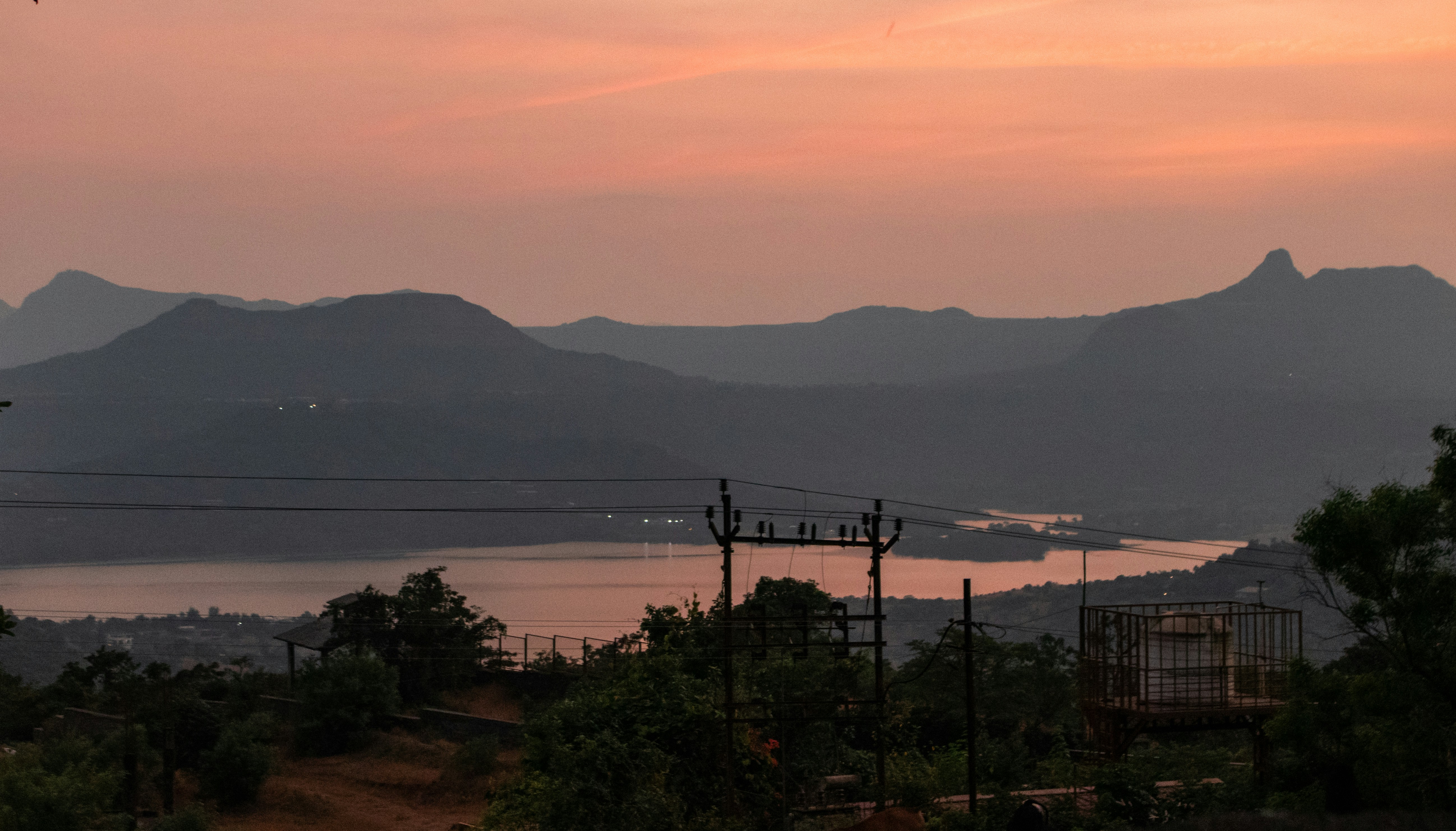 Silhouetted mountains against a pastel sunset sky with a lake reflecting warm hues and foreground foliage.