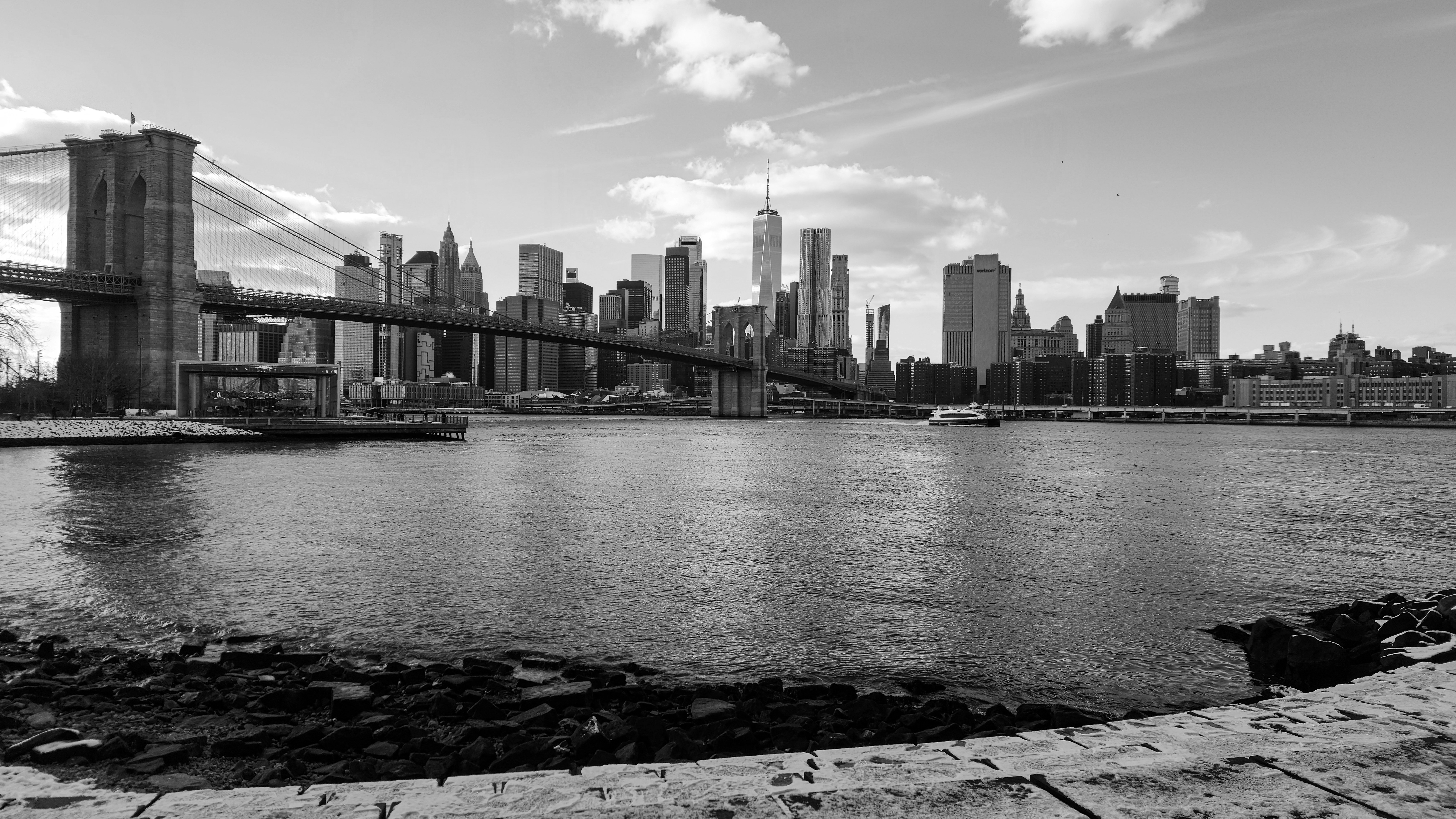 Brooklyn Bridge arches over the East River with the Manhattan skyline in the background, captured in black and white on a clear day.