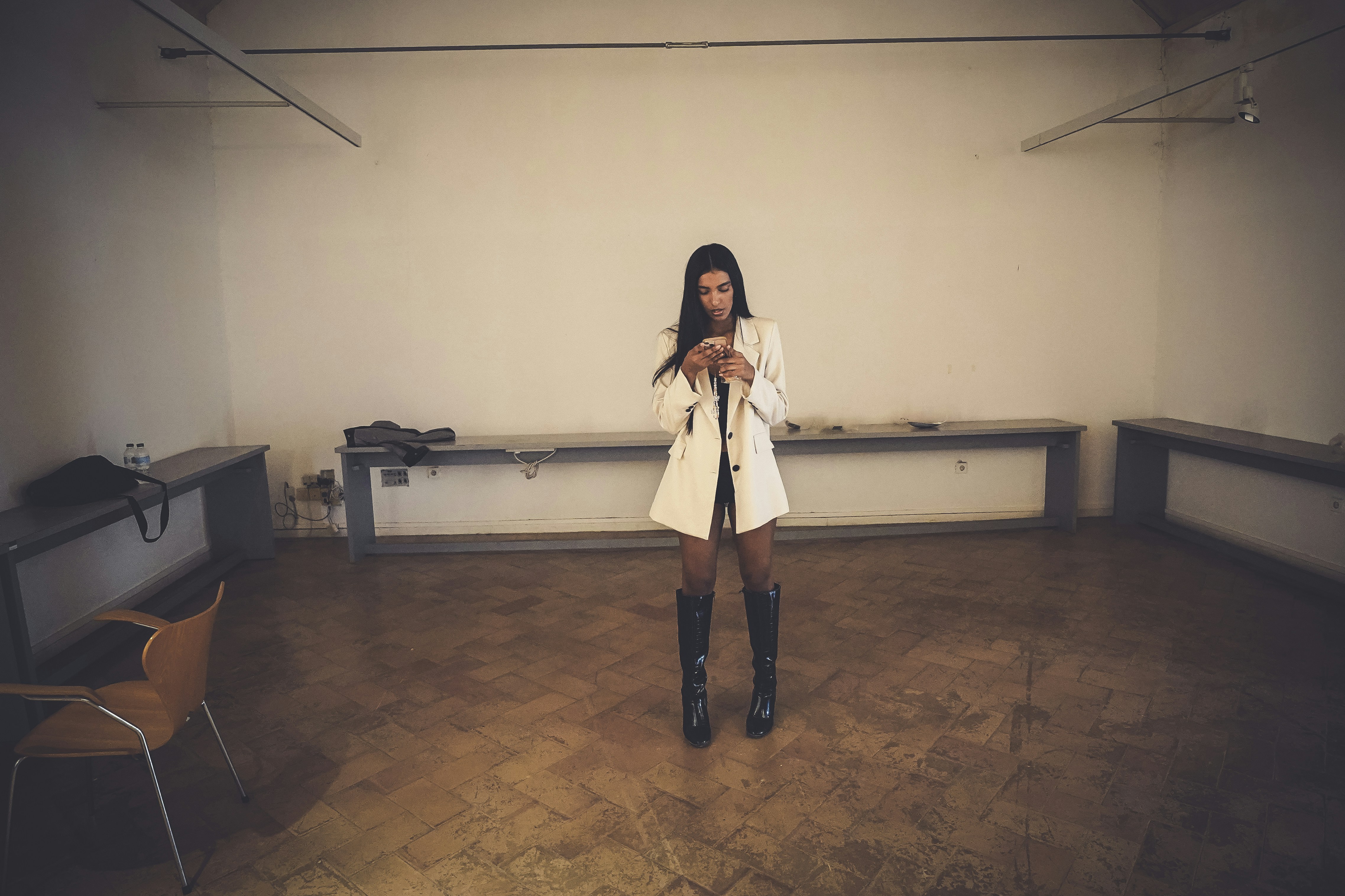 Woman in a white blazer and black boots focuses on her phone in a minimalist room with wooden floors and beige walls.