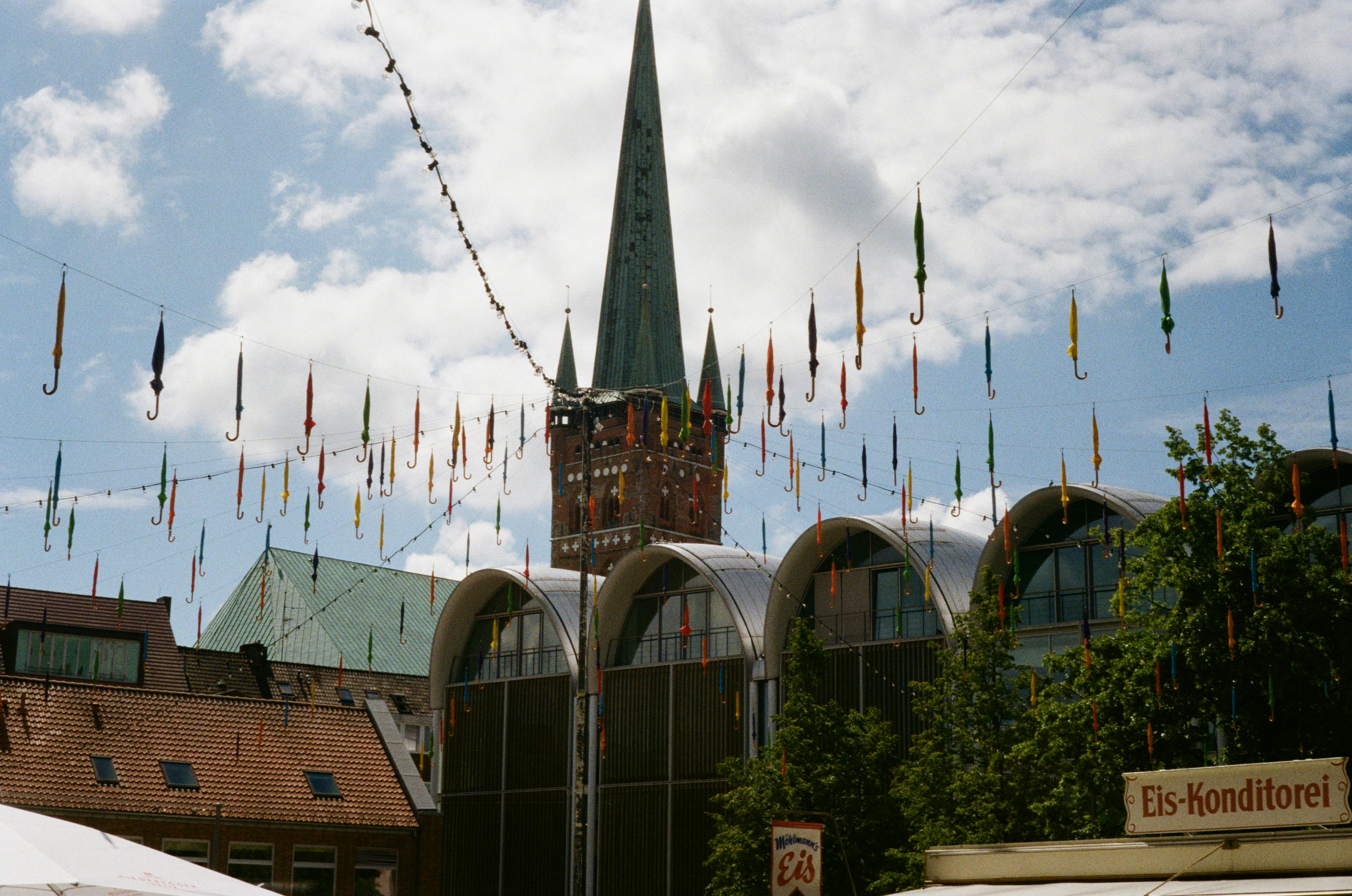 Tall church spire rises above arched building and colorful pennant strings under a partly cloudy sky.