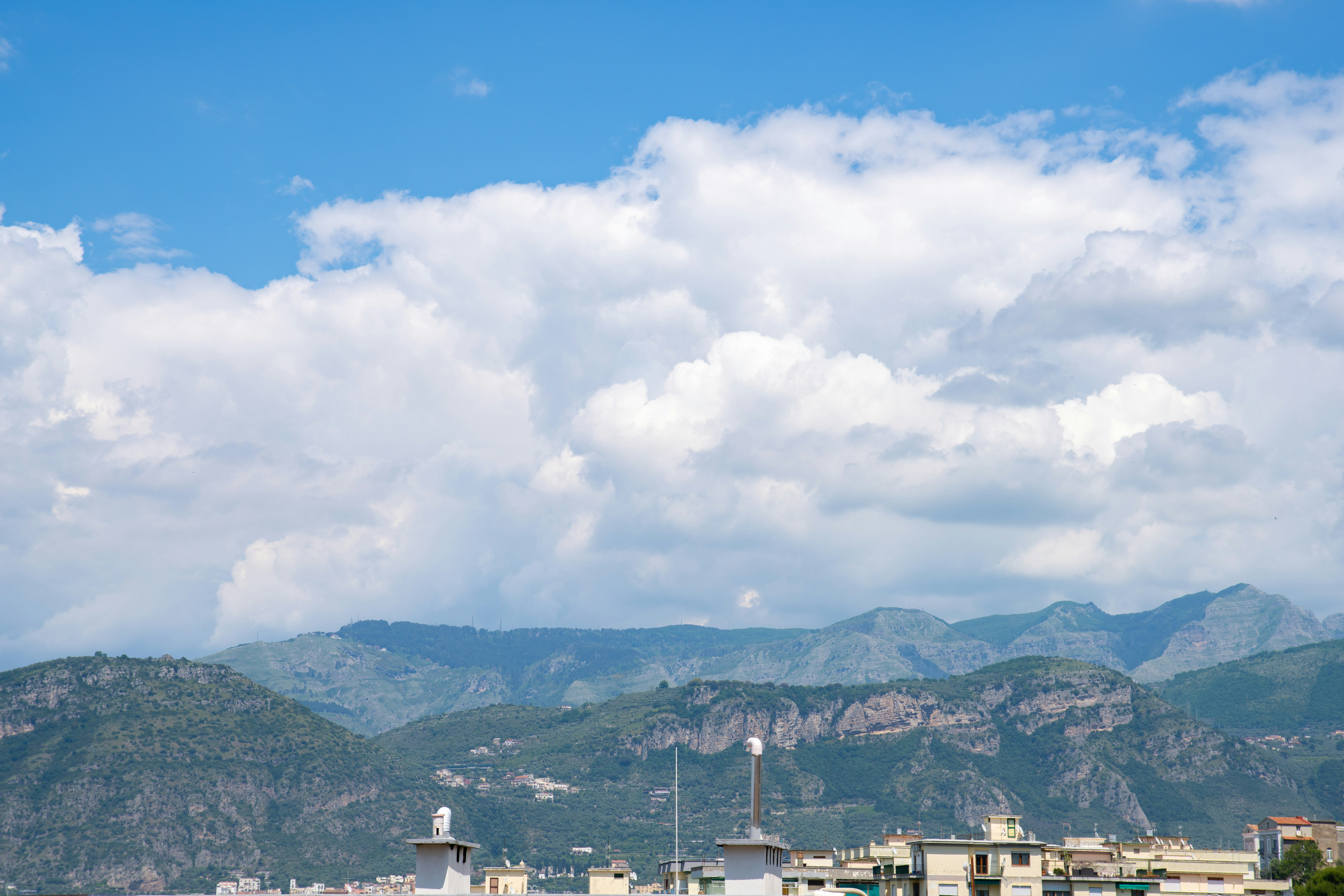 Mountain range under a vast sky with fluffy clouds, contrasting with urban rooftops in the foreground.