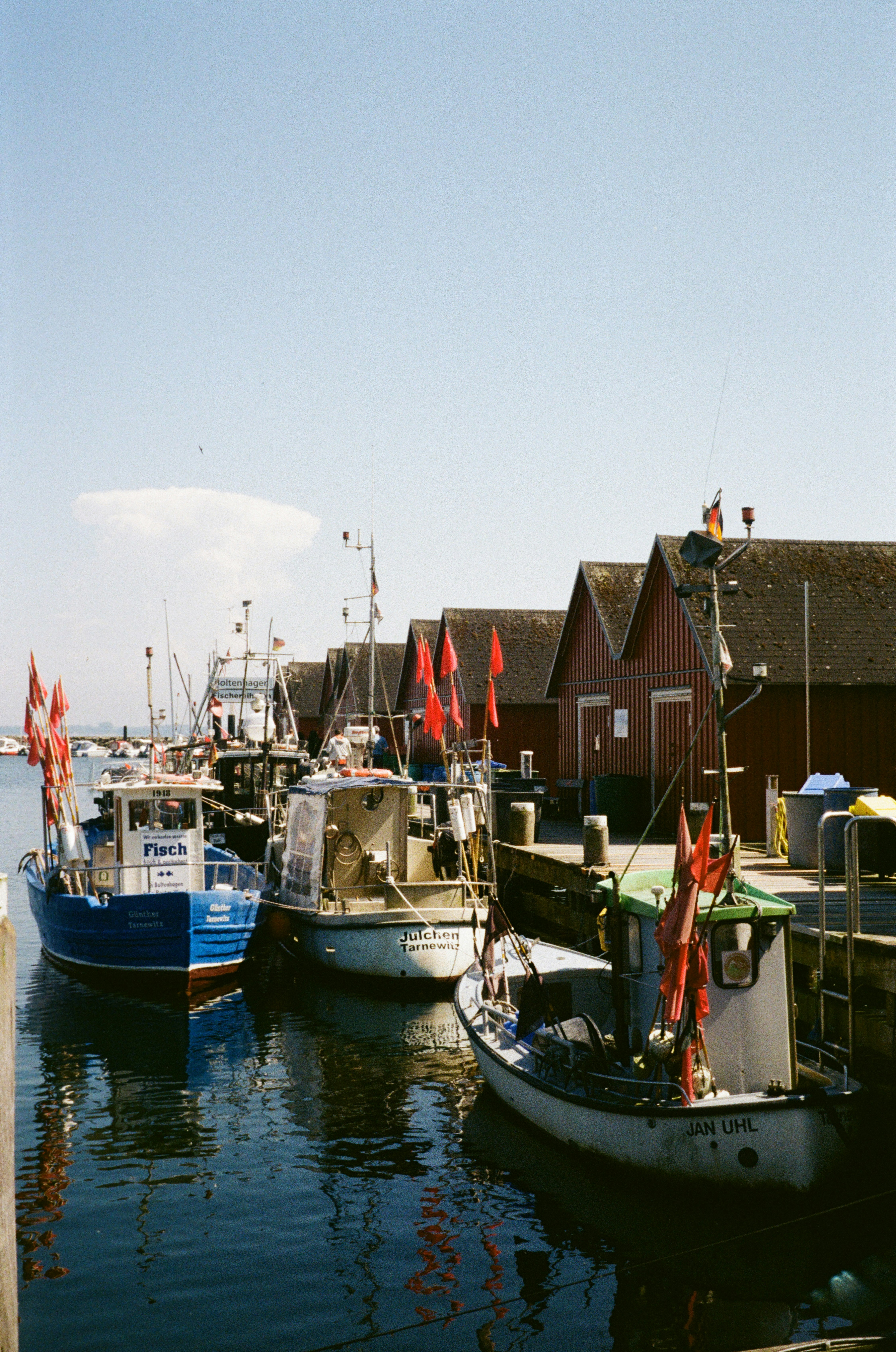 A harbor scene with colorful boats moored beside red wooden boathouses; reflections ripple across the calm water.