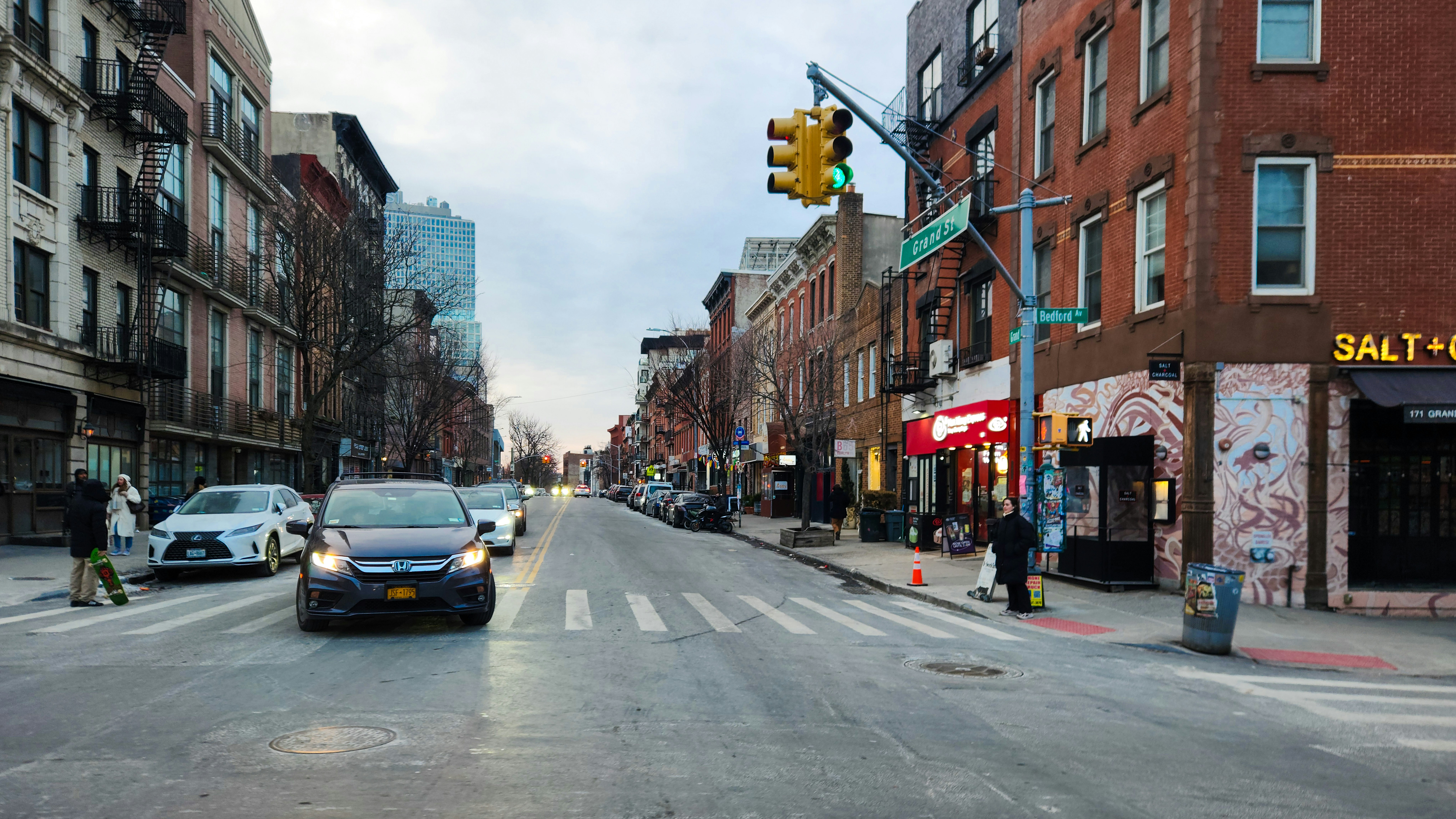 Cars and pedestrians navigate an urban street lined with red brick buildings and glowing storefronts.