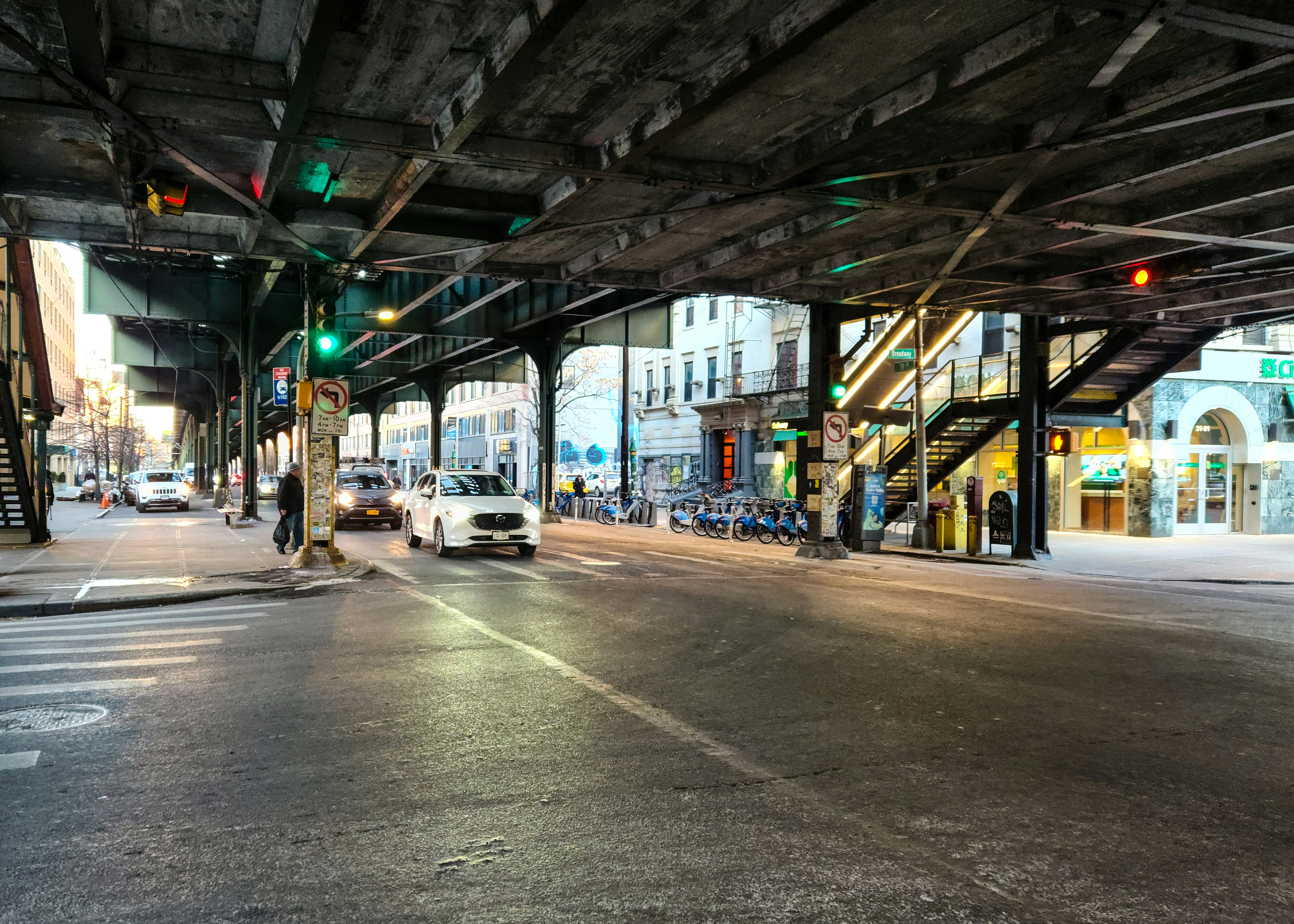 Cars pass beneath elevated subway tracks in a bustling city intersection at dusk.