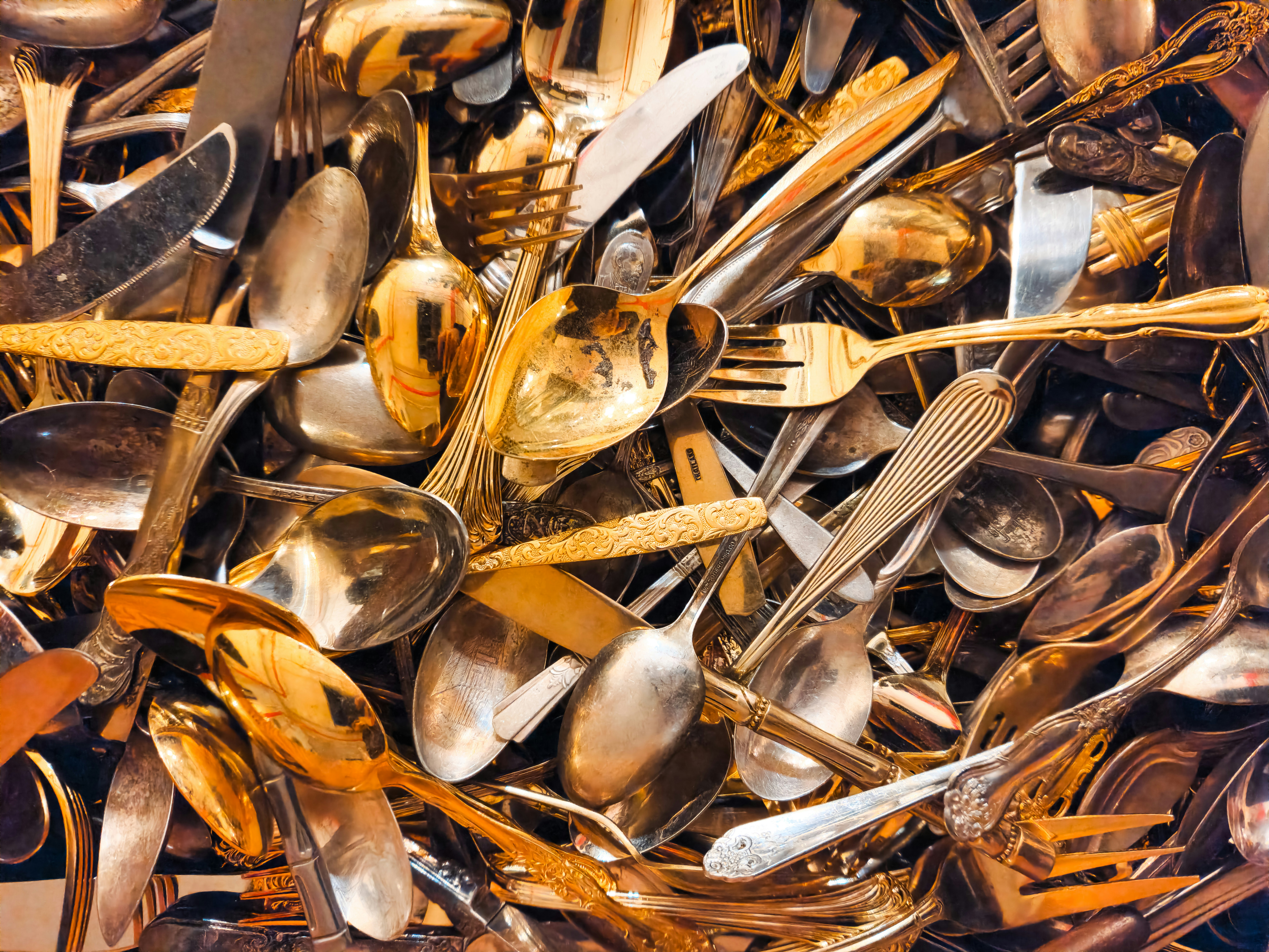 A collection of gold forks and spoons piled up in a box, showcasing their elegant design and luxurious appeal. These pieces of cutlery, often used for special occasions or high-end dining, feature intricate detailing and a gleaming finish. The box presents a refined and organized way to store these precious utensils, which can add a touch of sophistication and opulence to any dining setting. The gold hue gives the silverware a timeless and collectible charm. | A pile of old metal spoons and forks