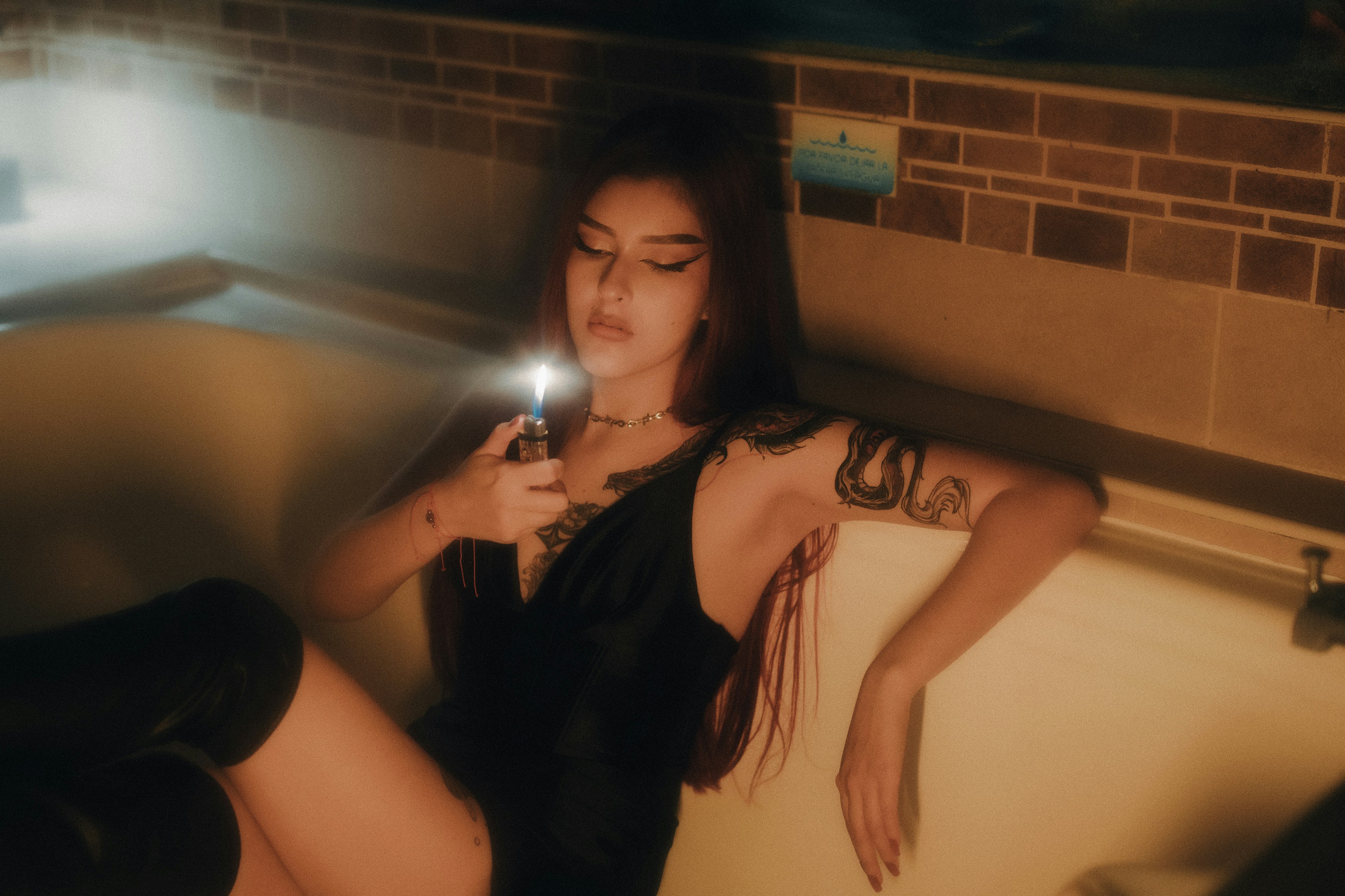Woman holding a lighter in a bathtub, surrounded by warm ambient light. Intricate tattoos and soft shadows create an intimate, contemplative atmosphere.