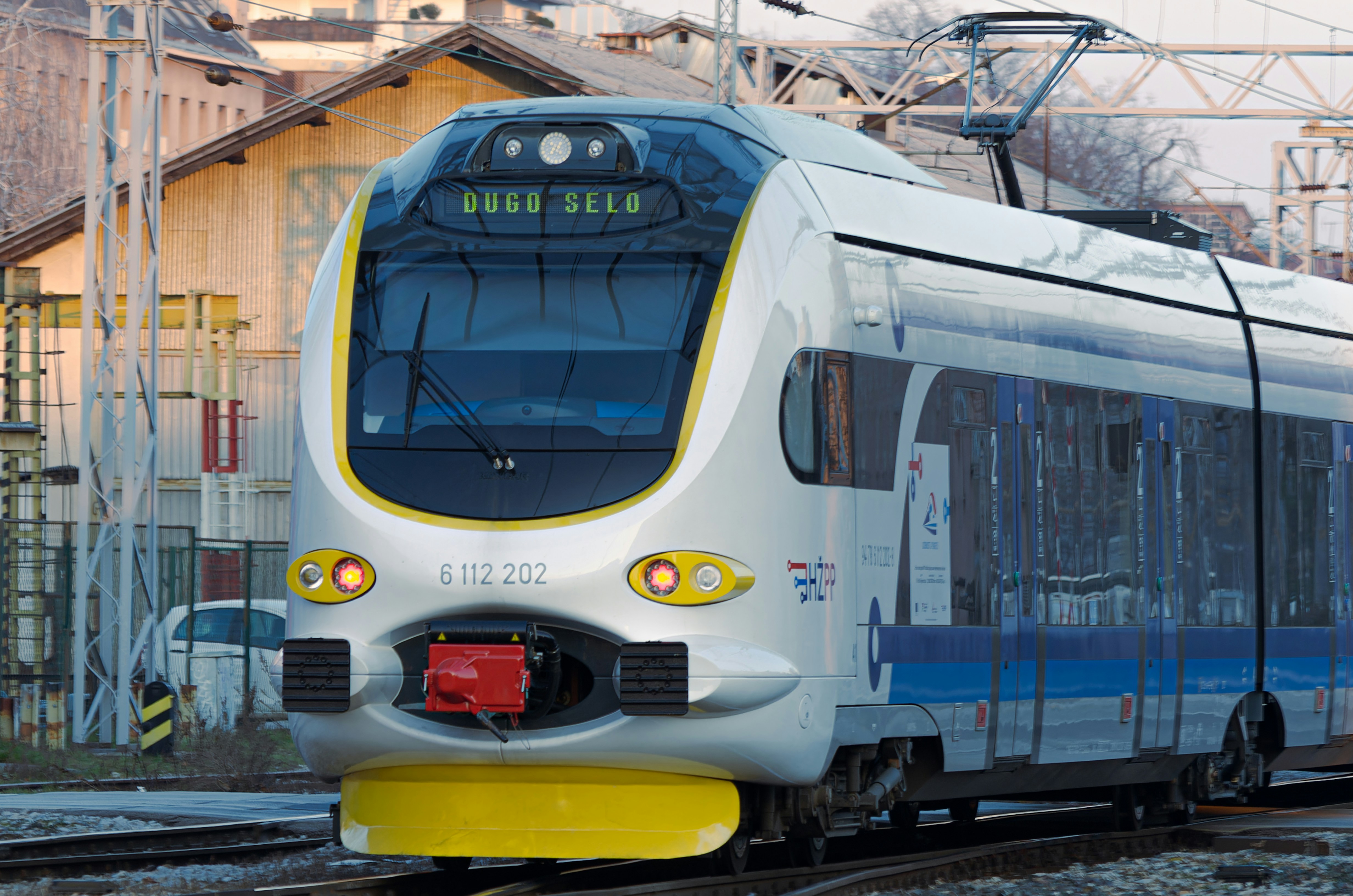 Sleek train with blue and white exterior approaches a city station, contrasting with industrial surroundings.