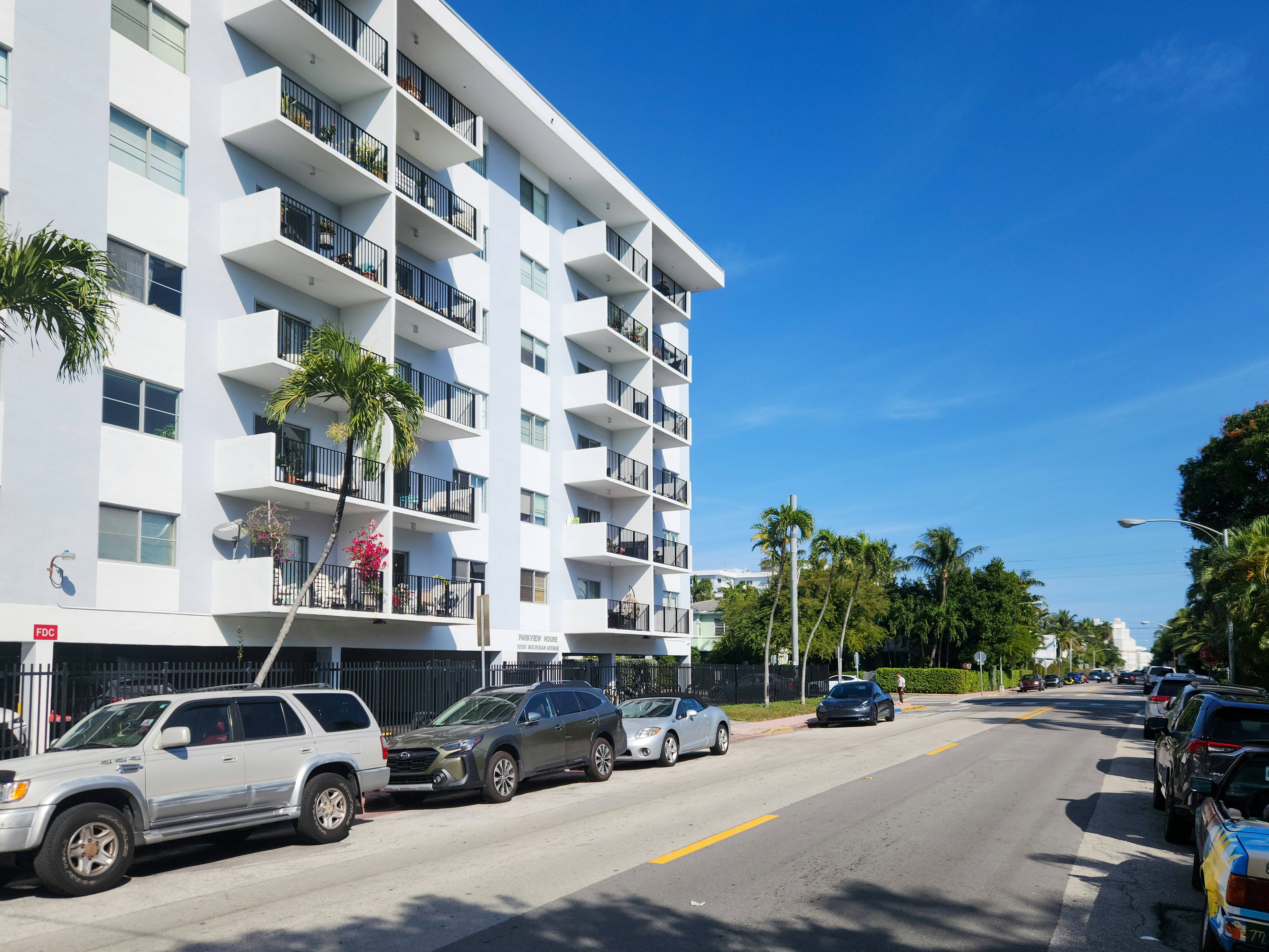 Modern apartment building with white facade and balconies, contrasted by green palm trees and blue sky on a sunny street.