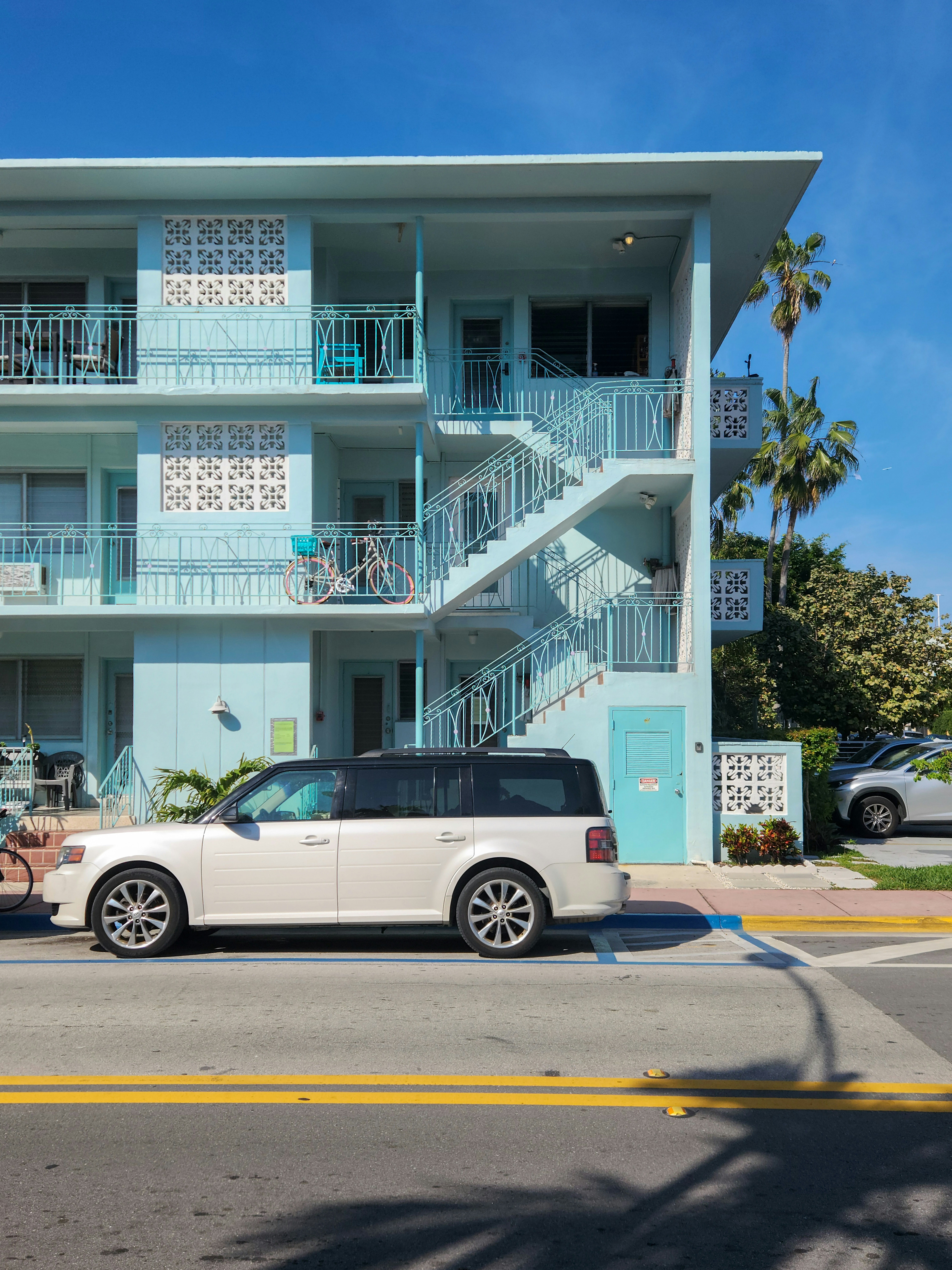 Light blue Art Deco building with geometric patterns and balconies, framed by palm trees under a clear sky.