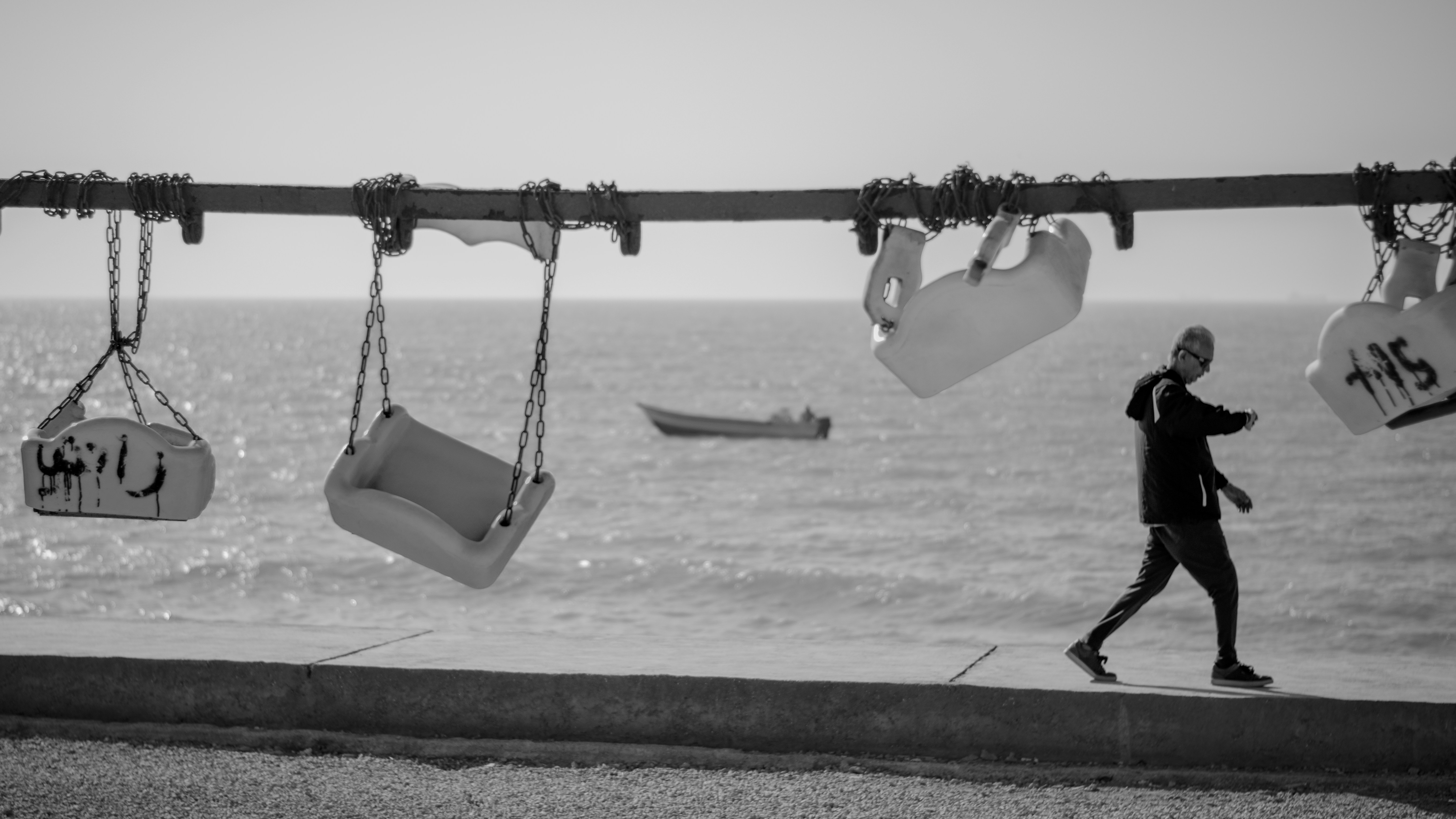 Black and white scene of a person walking past hanging swings by the sea, with a lone boat in the distance.