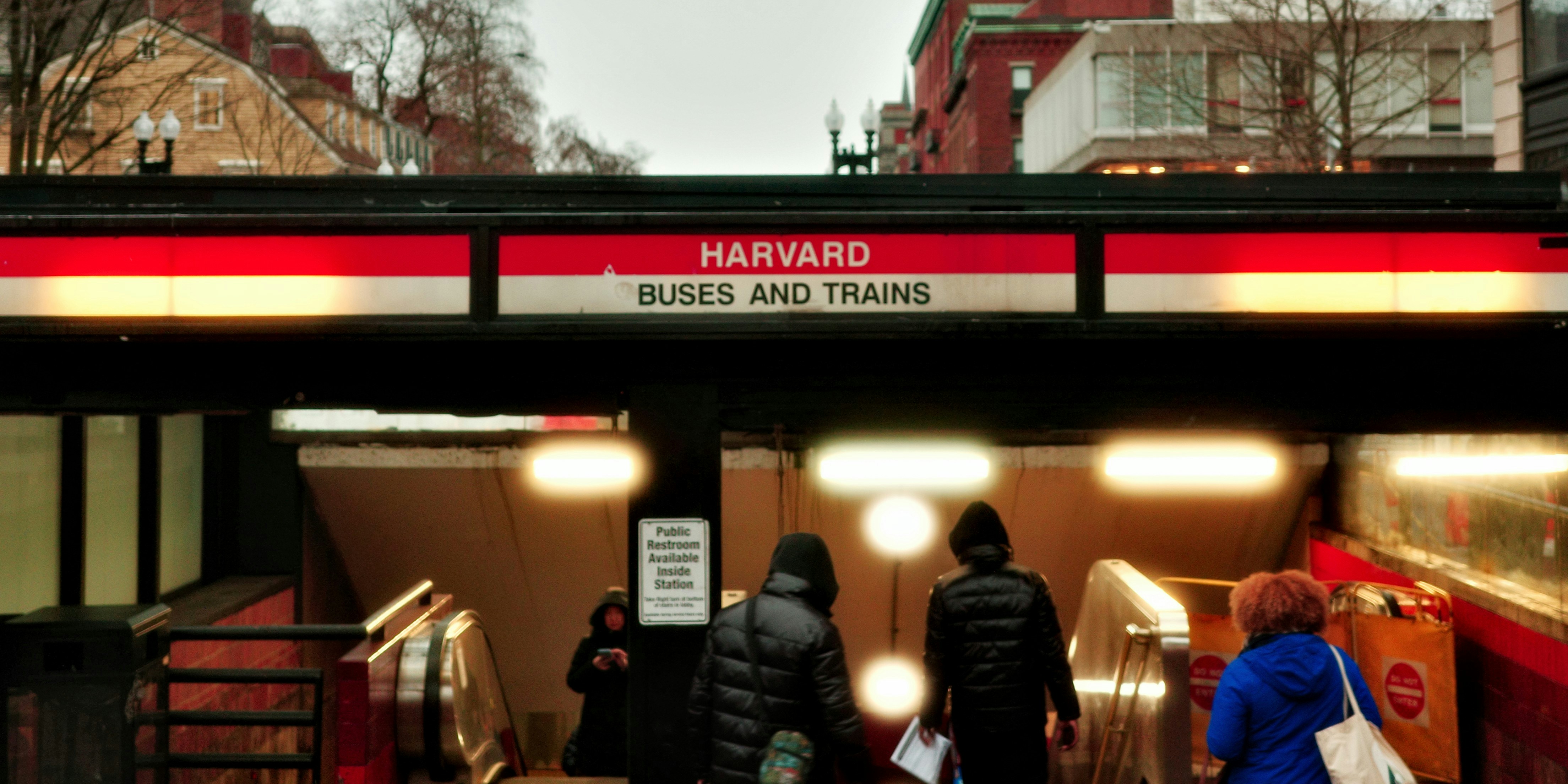 Commuters enter and exit the Harvard Square subway station under a cloudy sky.