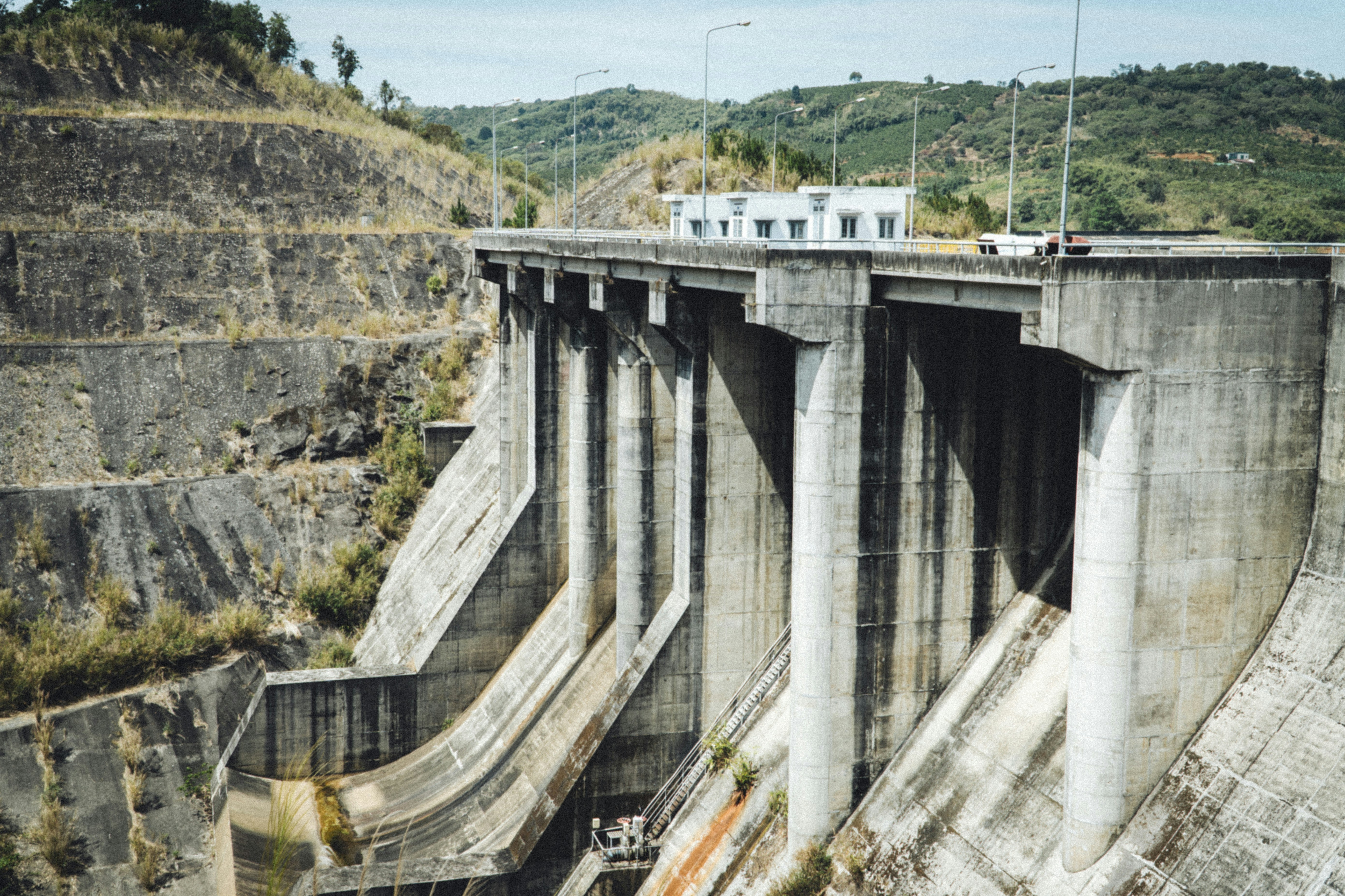 Massive concrete dam with spillways set against a backdrop of rolling hills and greenery.