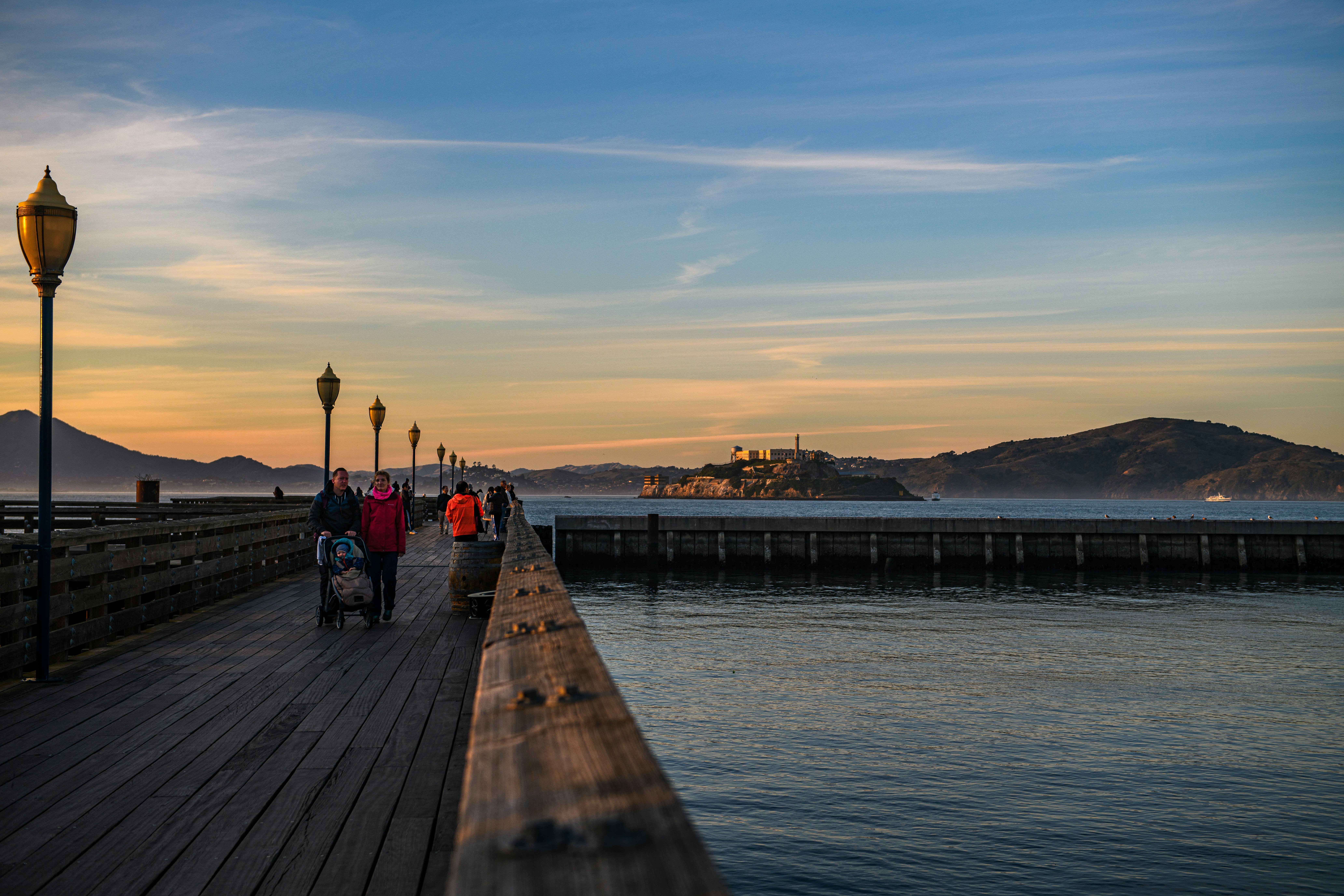 Wooden pier stretching into a calm sea under a sunset sky, with people walking and distant hills on the horizon.
