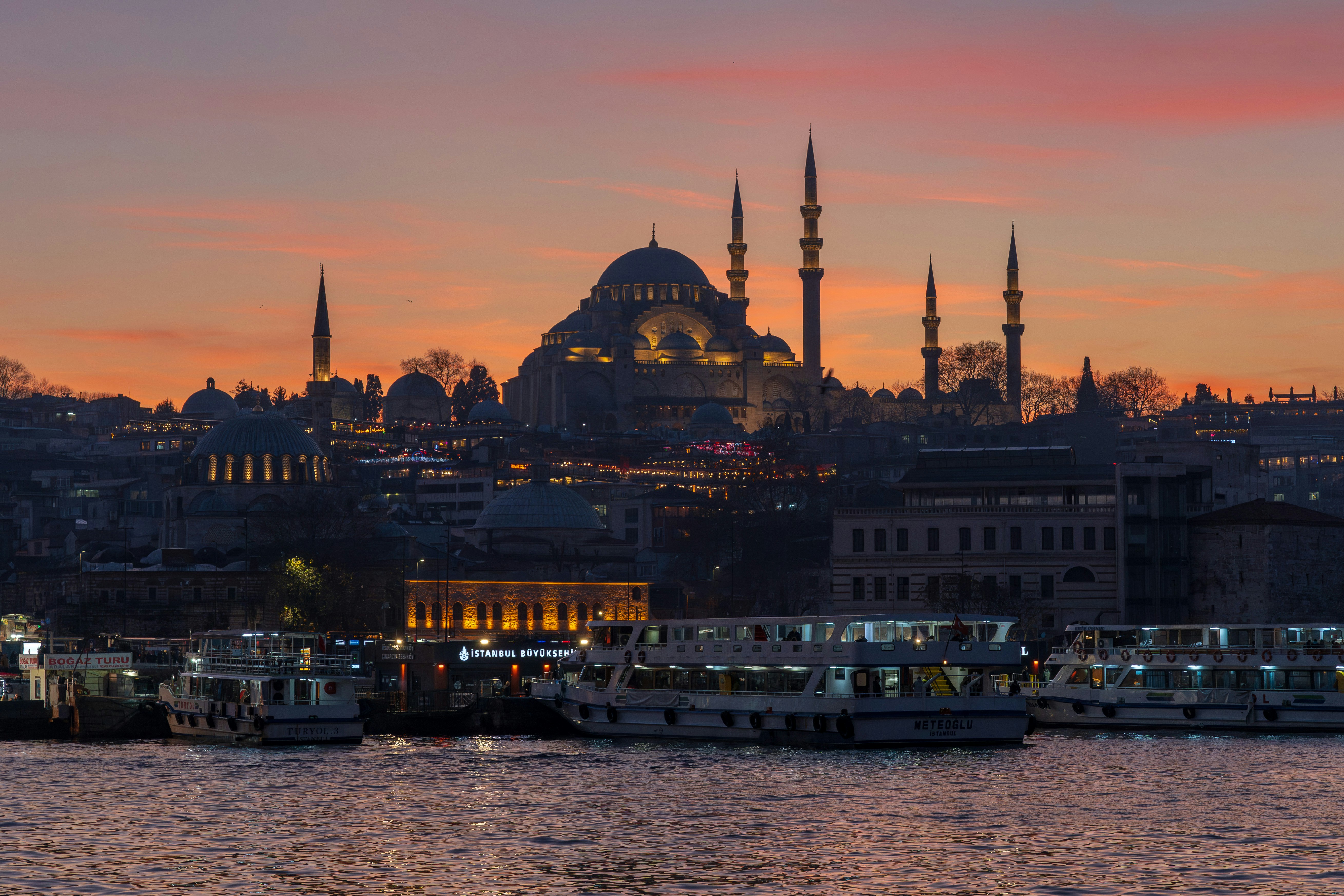 Silhouette of a mosque at sunset with vibrant skies and ferry boats in the foreground.