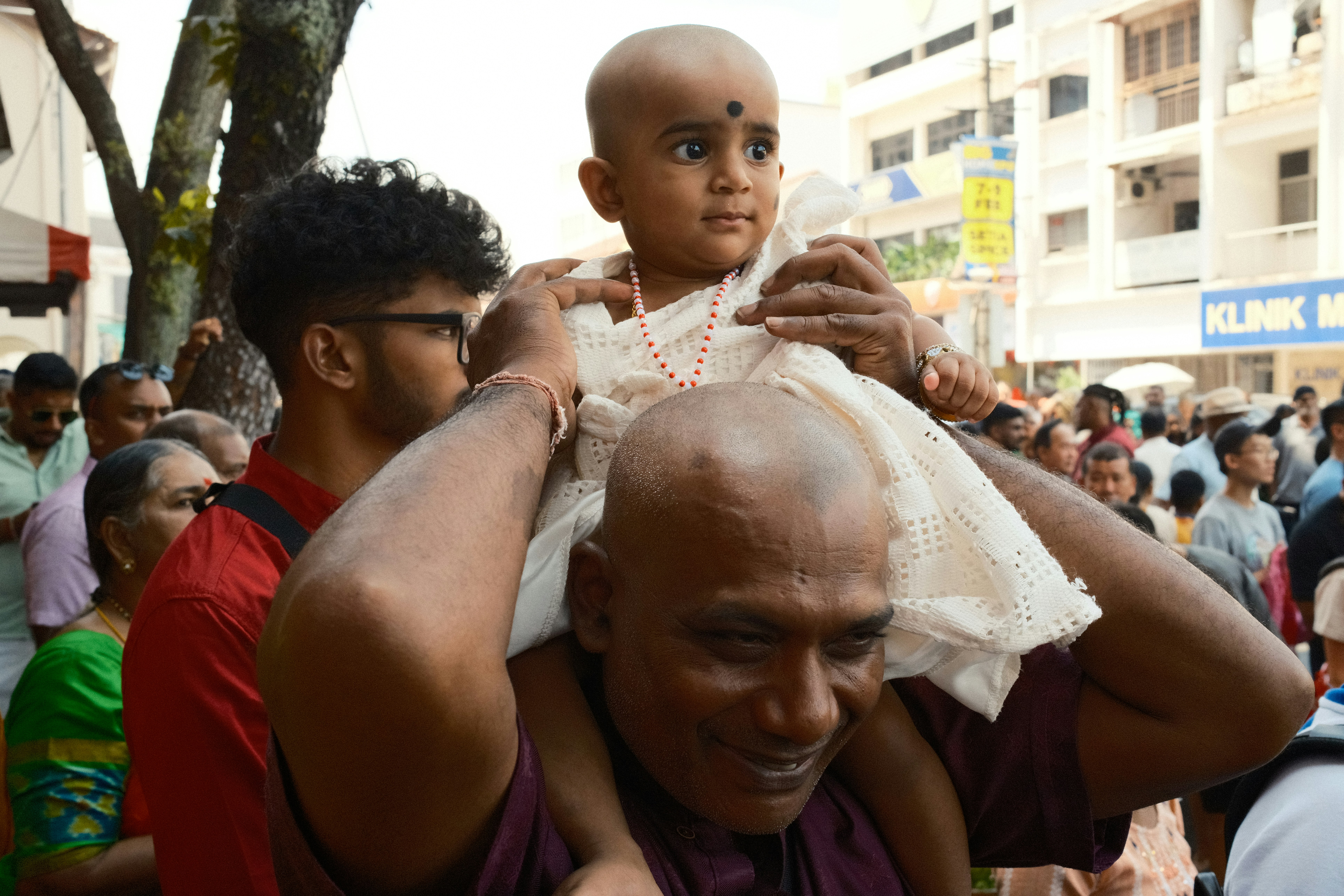 Child in white attire sitting on an adult's shoulders amidst a lively crowd in warm lighting.