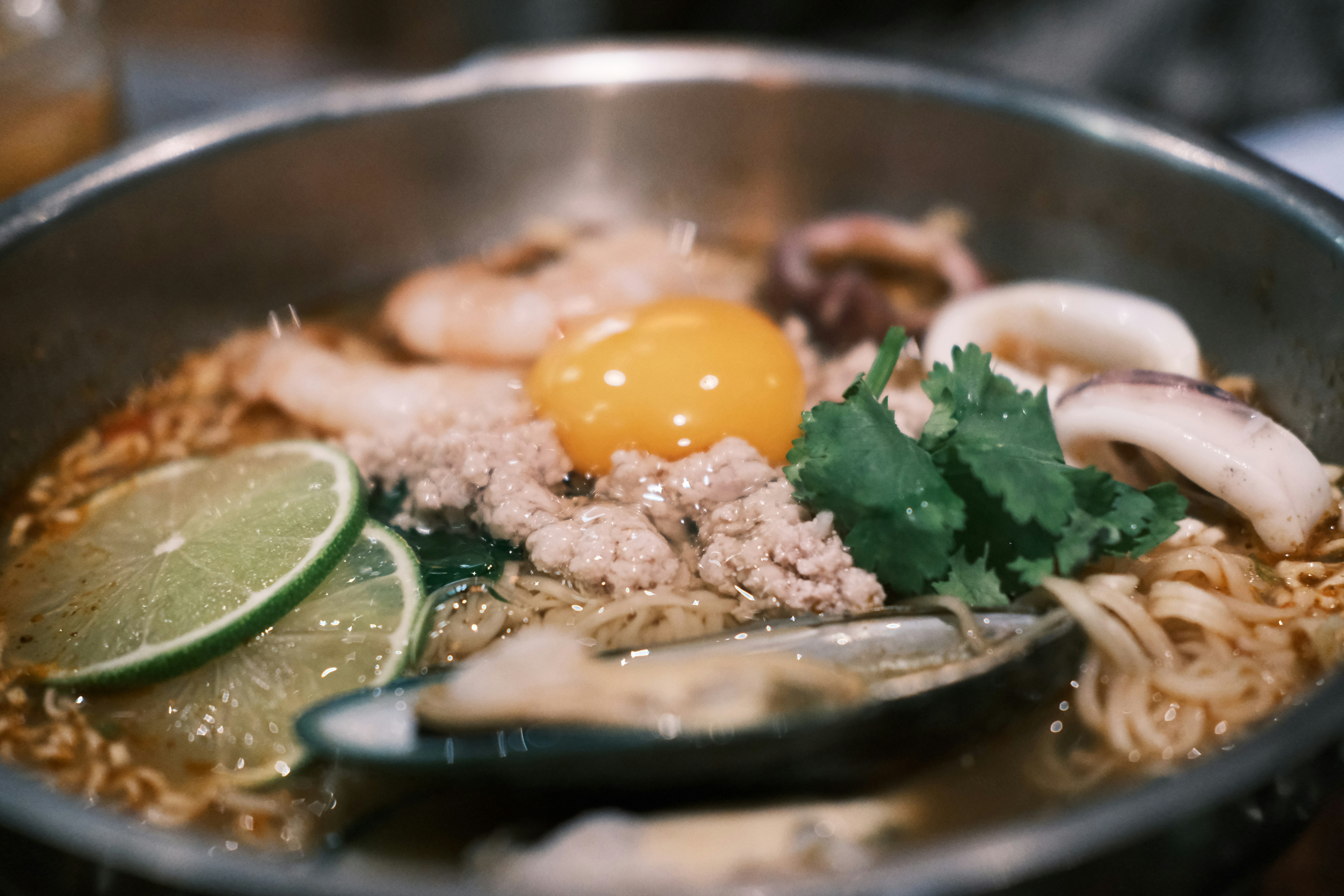 Seafood ramen topped with lime, egg yolk, cilantro, shrimp, and squid rings in warm lighting.