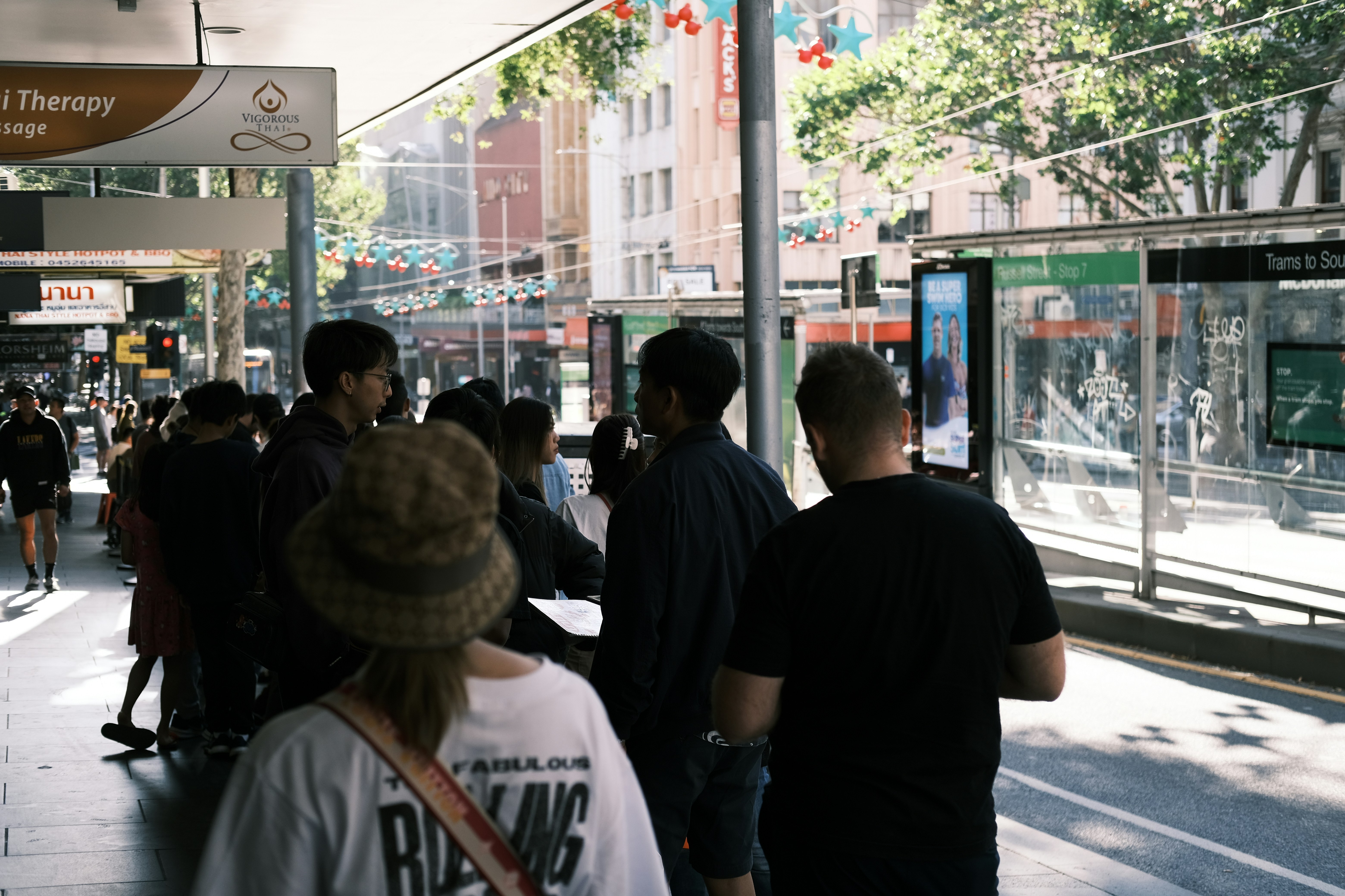 Crowd of people waiting at a tram stop under a canopy, with sunlight streaming through trees and colorful urban signage.
