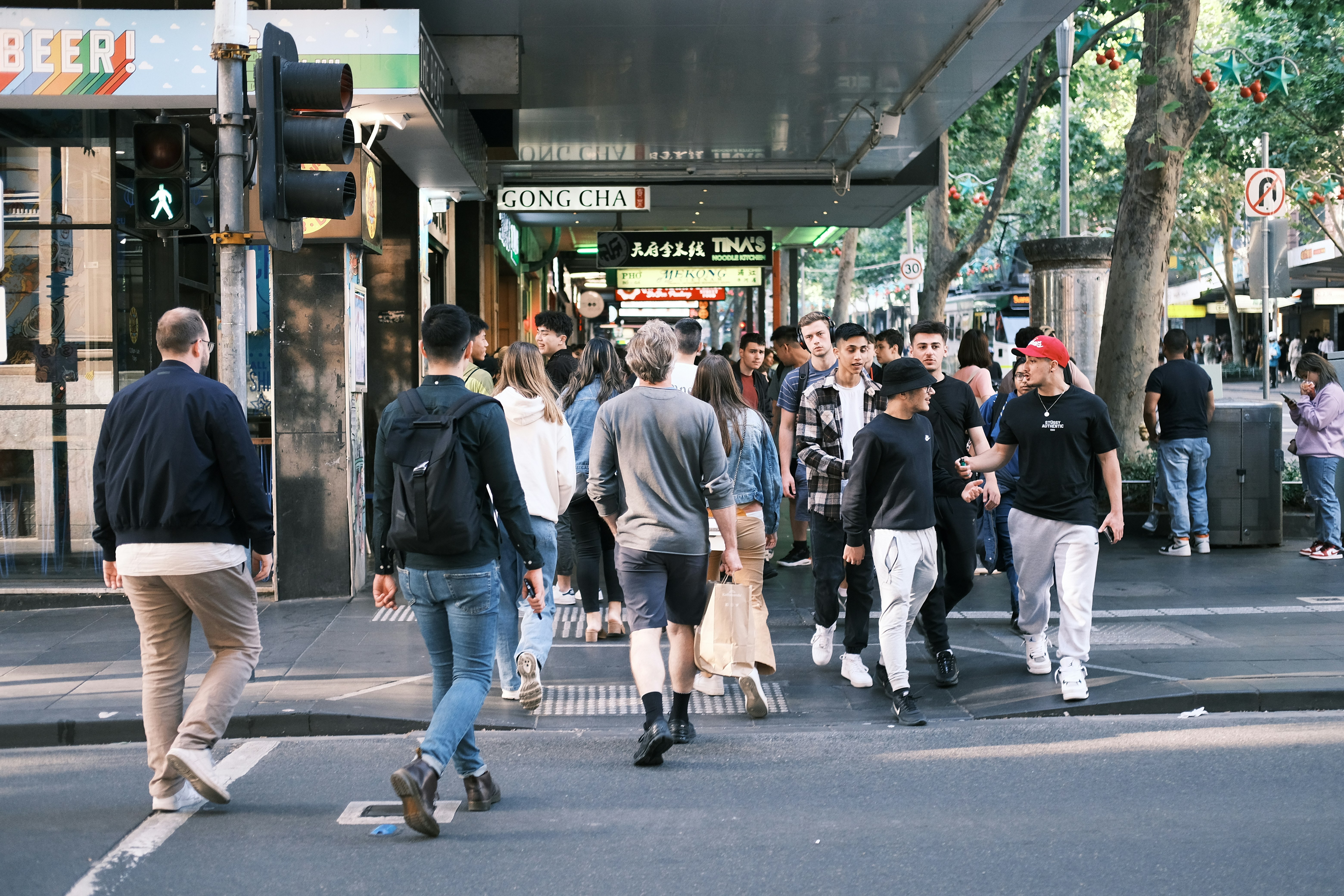 Pedestrians fill a vibrant city crosswalk beneath storefront awnings and trees.
