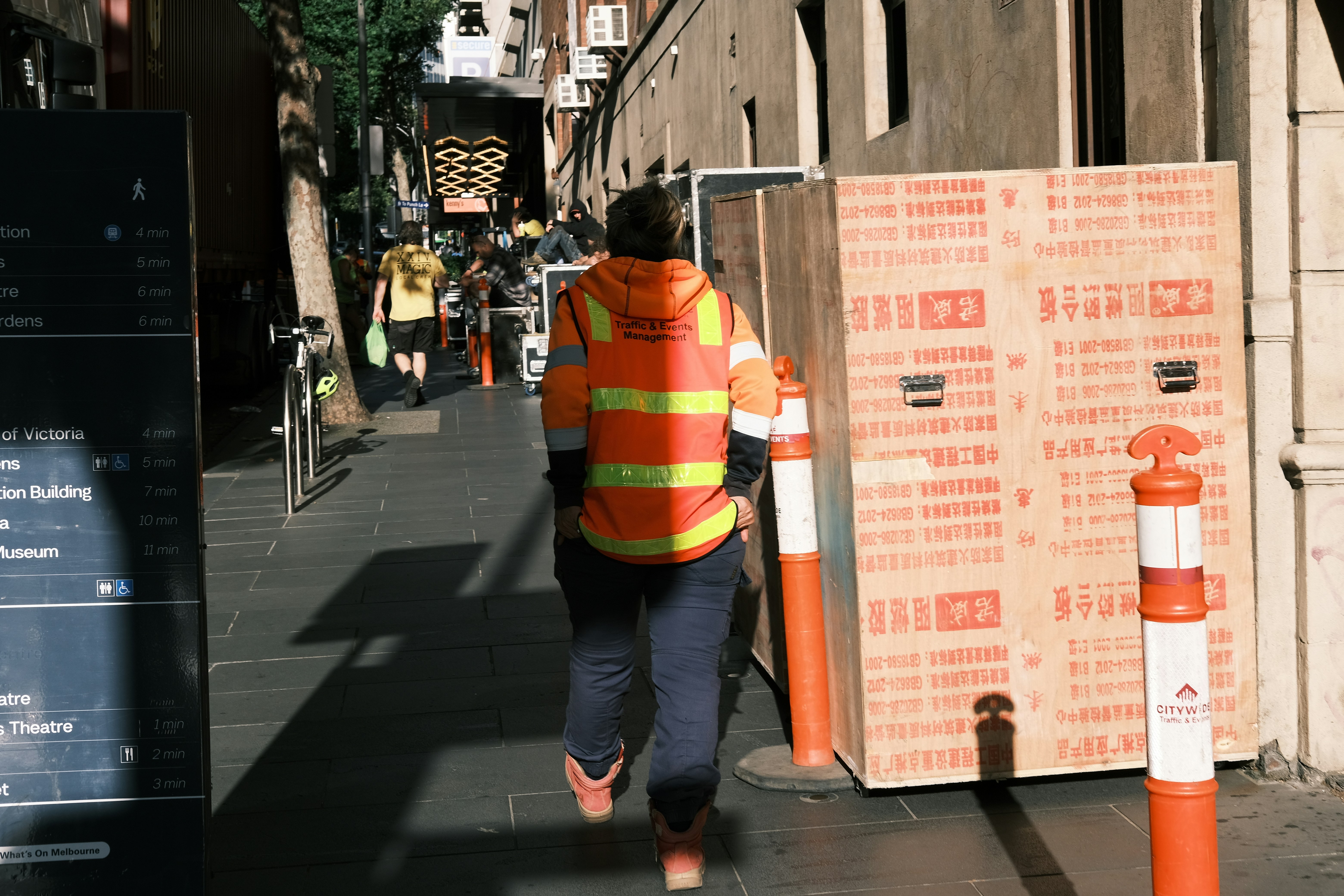 Worker in a bright orange vest walks along a sunlit city sidewalk surrounded by construction elements.